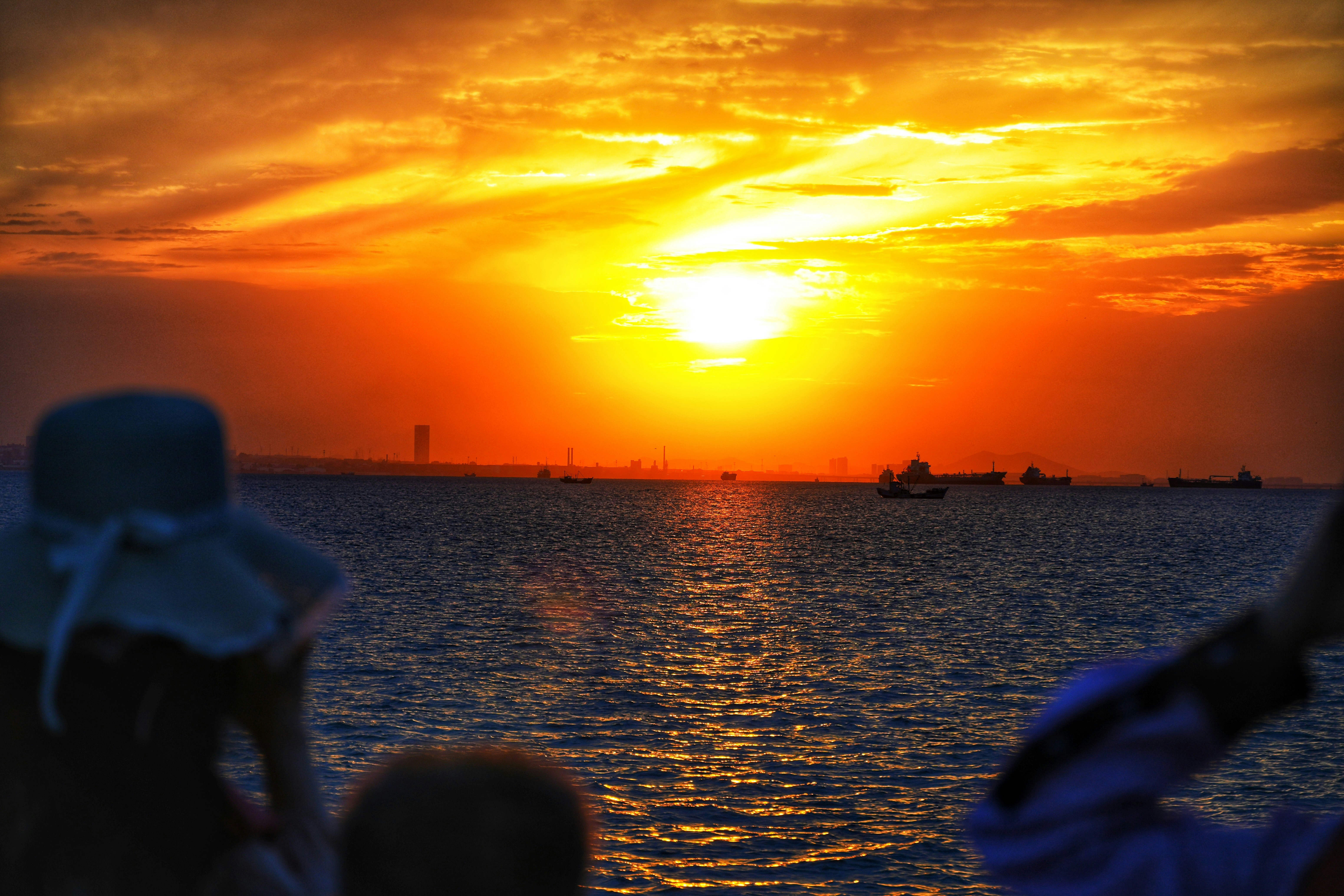 Golden sunset over calm water with silhouettes of boats on the horizon; foreground figures blur as the scene captures a tranquil harbor moment.