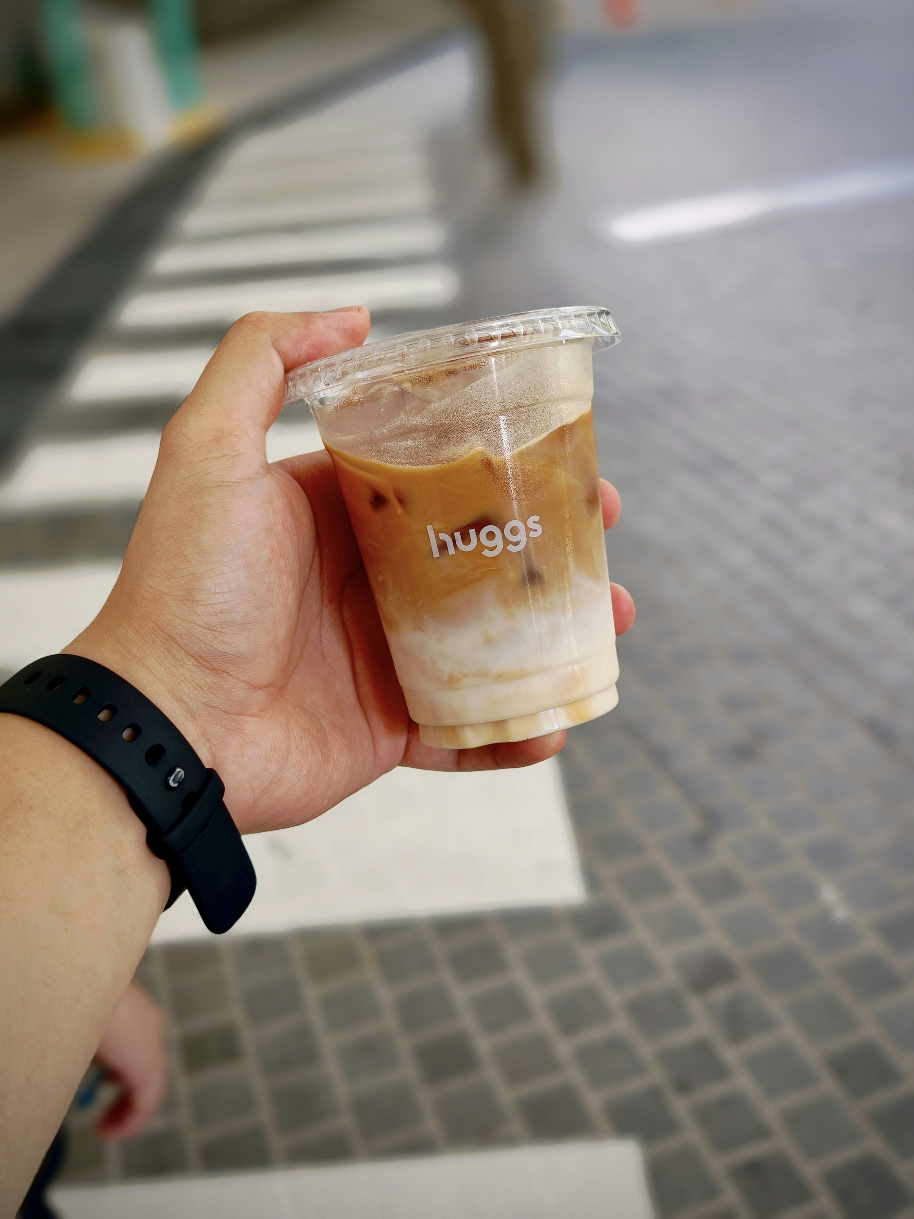 Cold coffee cup with logo hugged by a hand; blurred urban sidewalk backdrop and crosswalk lines in the distance.