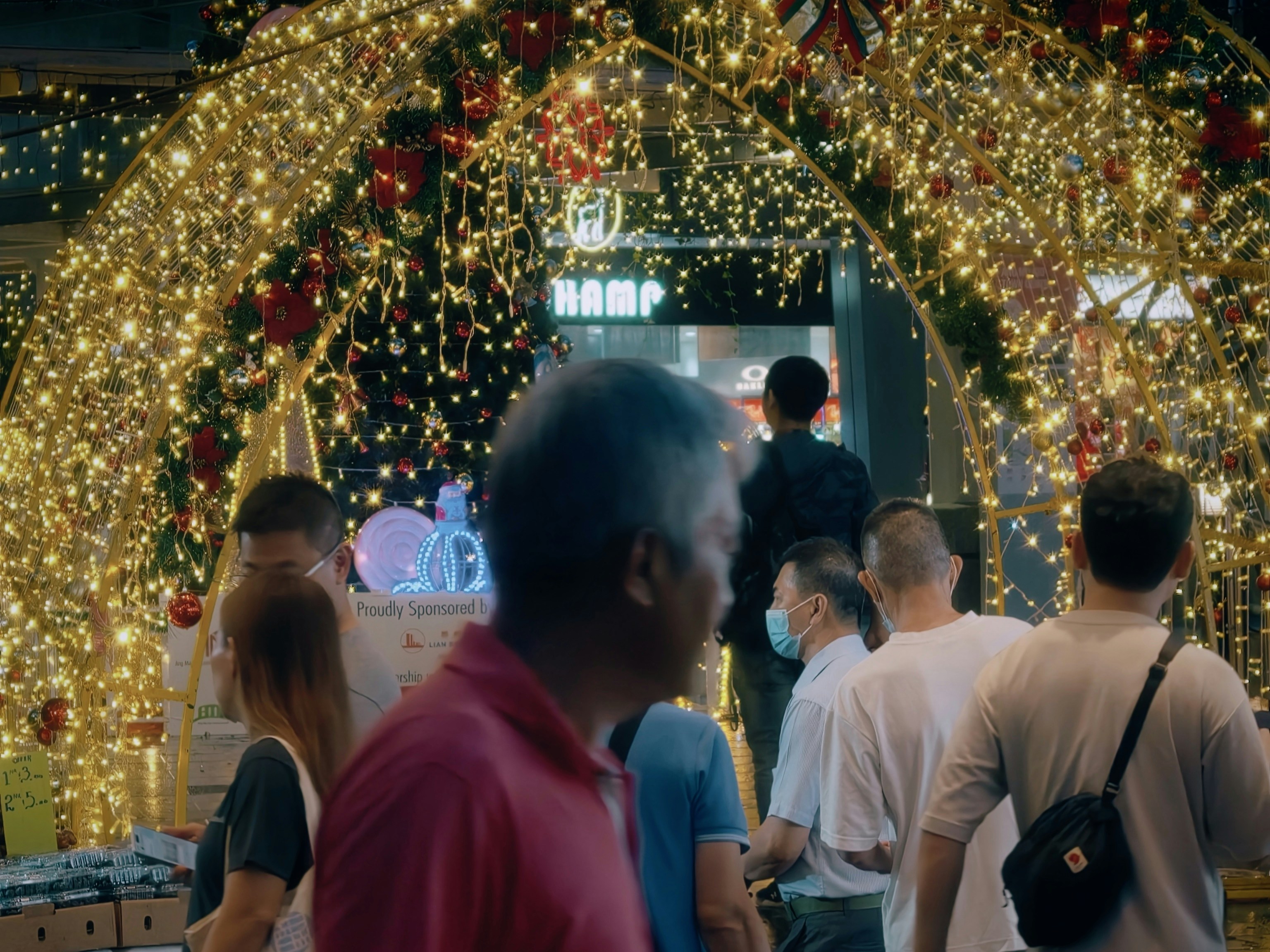 Vivid festive arches of string lights frame a crowded indoor scene with pedestrians, some wearing masks, creating a warm glow and sense of movement.