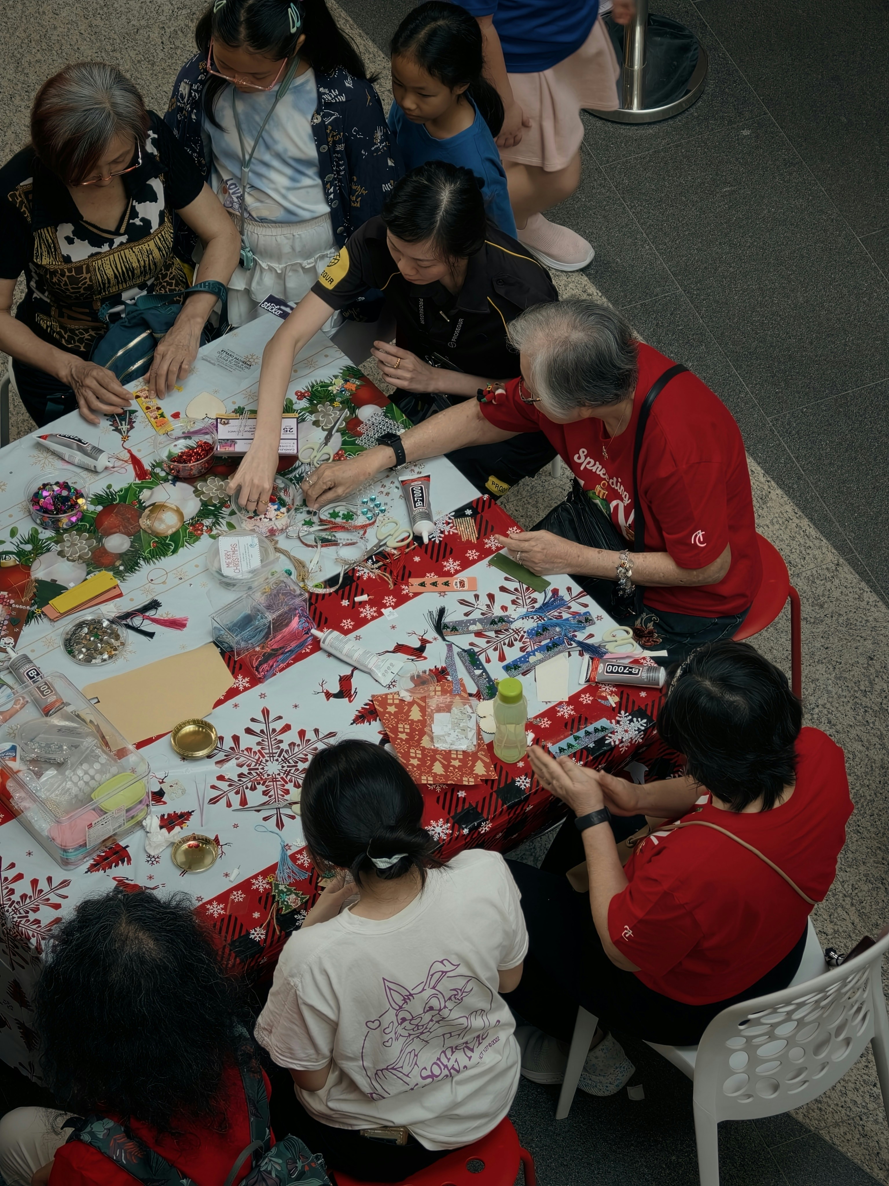 Bright craft table with red and white patterns as a group, including adults and children shaping decorations and arranging supplies.