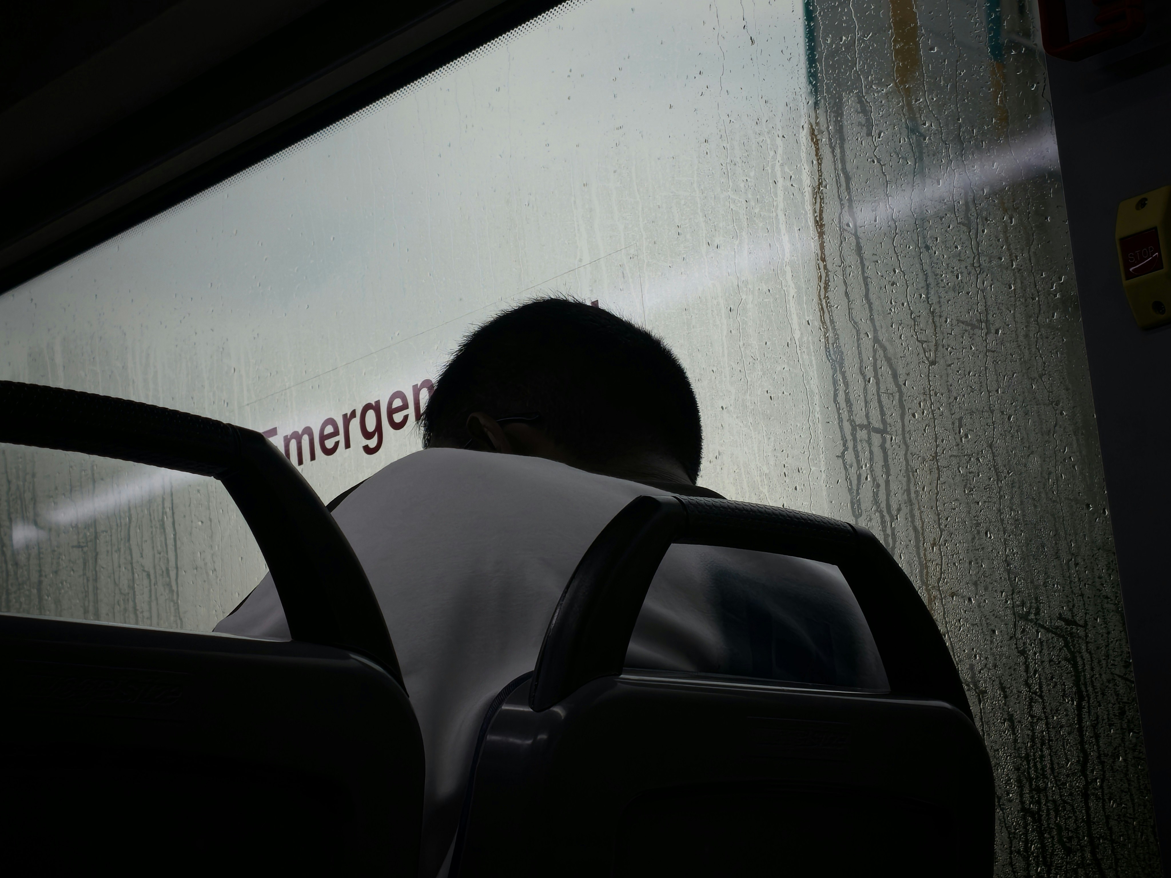 A dark, inside-of-bus scene with a person leaning forward against a seat; light from the window reveals a misty, blurred exterior and a partial red emergency sign. This is a photograph, not a drawing.