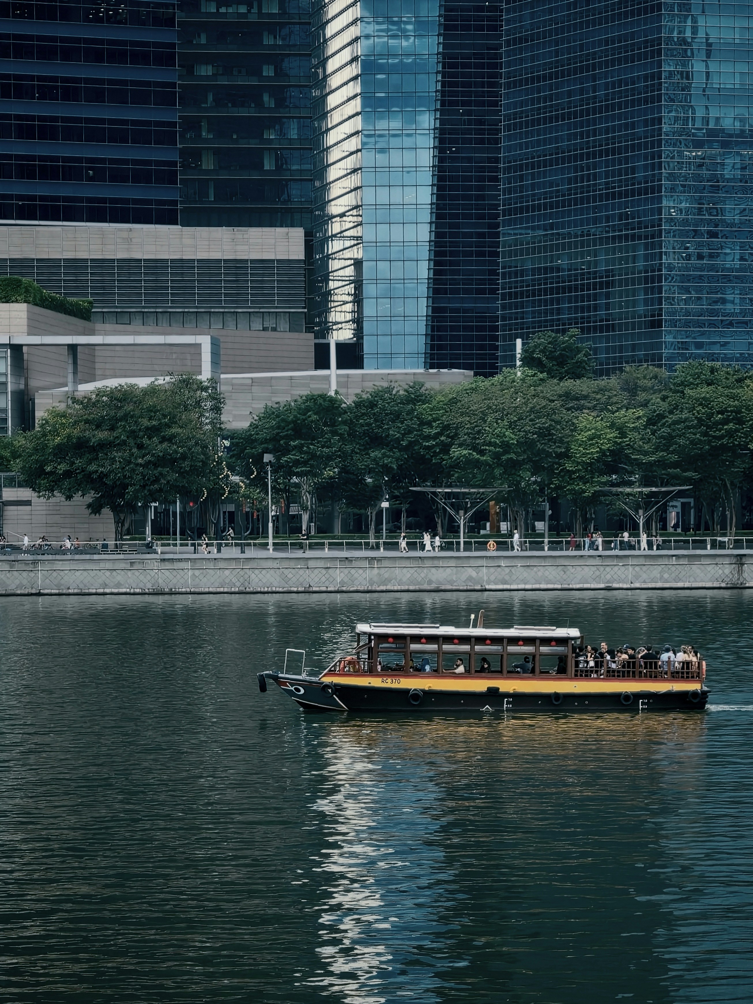 Brown passenger boat glides along a river with a modern skyline and green trees in the background.