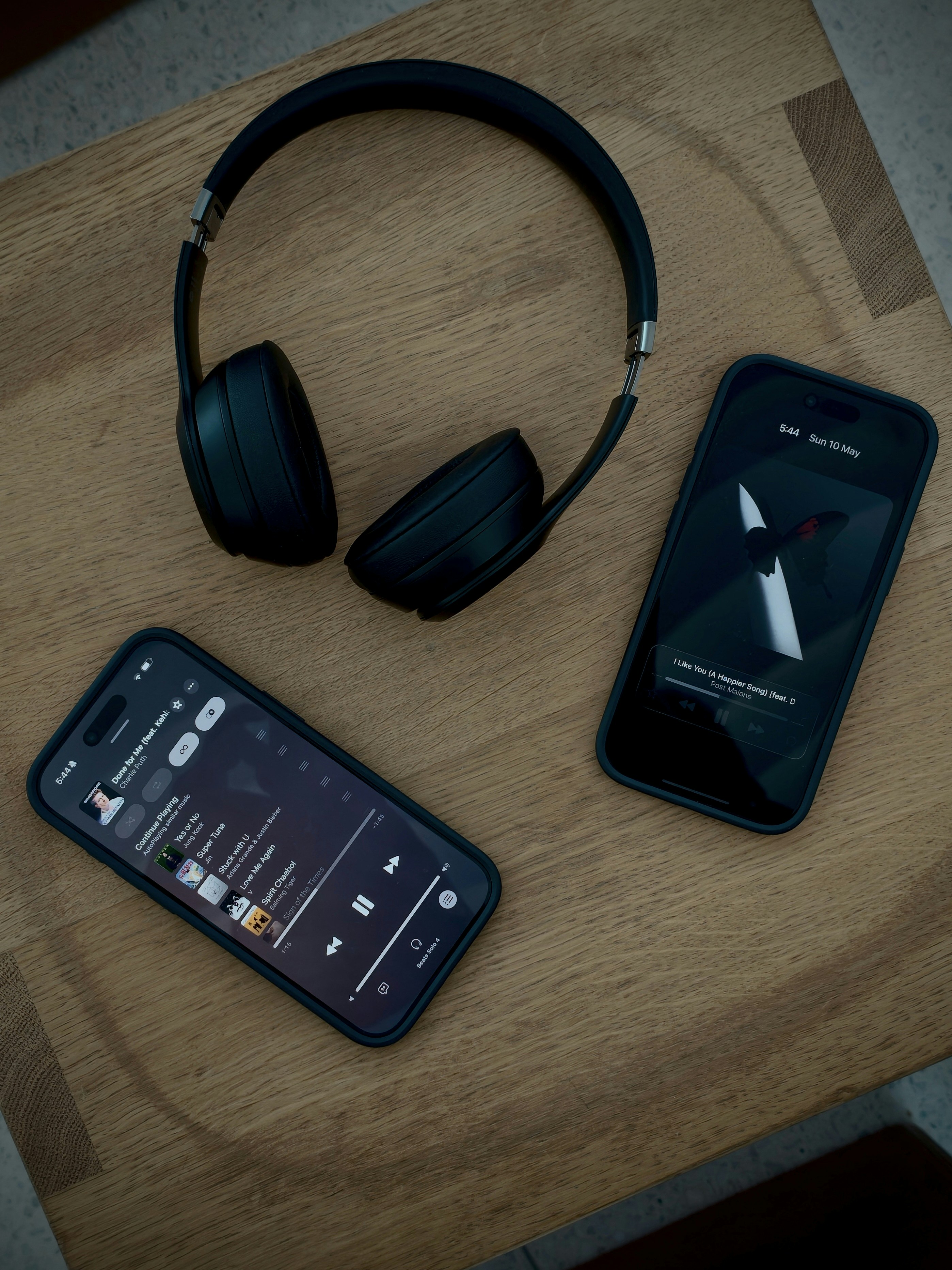 Two smartphones and a pair of over-ear headphones rest on a wooden desk, with a moody, low-light ambiance and subtle screen glow on the devices.