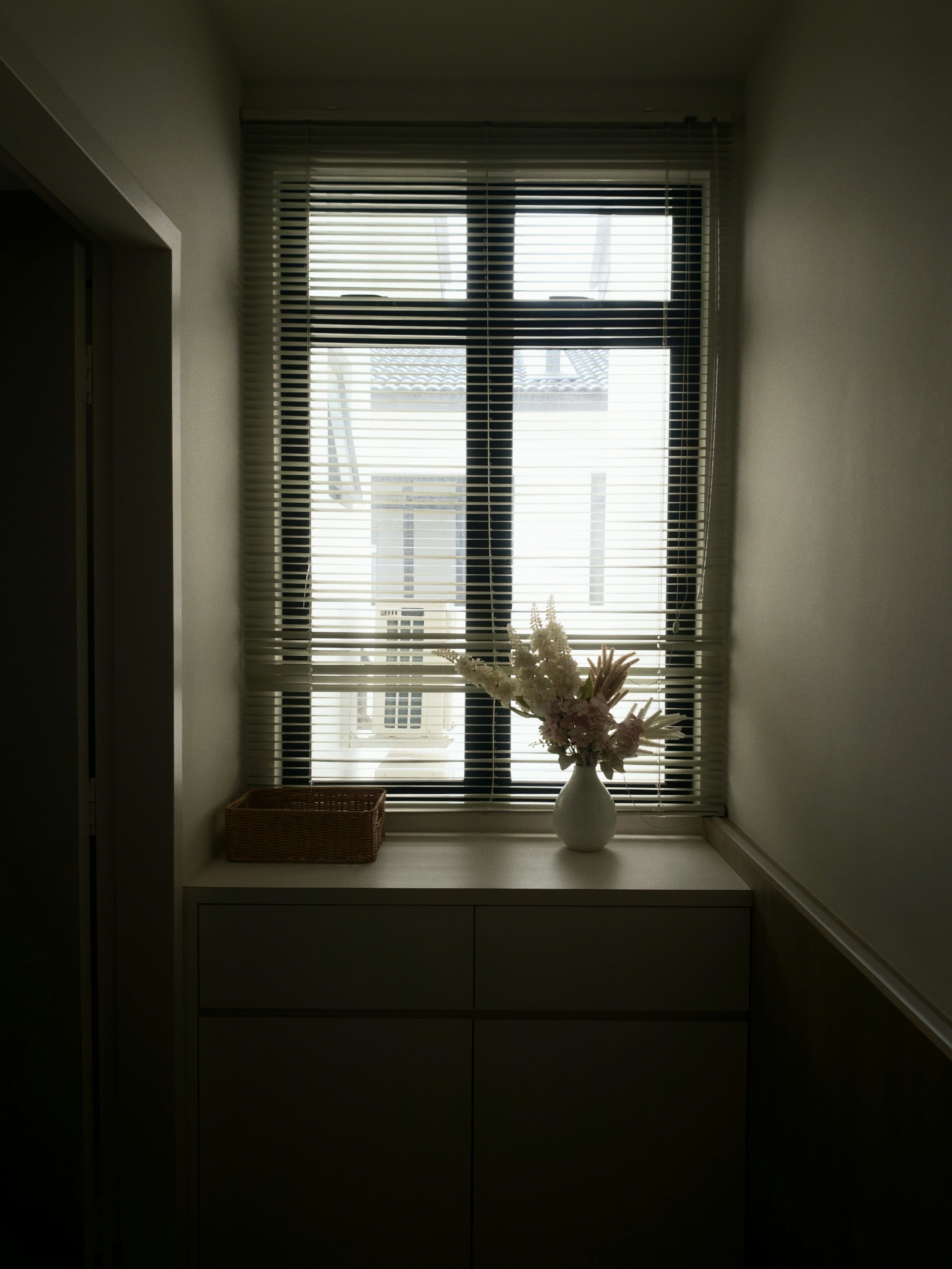 Sunlit window with horizontal blinds filtering pale daylight, a white vase of dried flowers sits on a cabinet beneath, a small basket to the left, creating a calm interior still life.