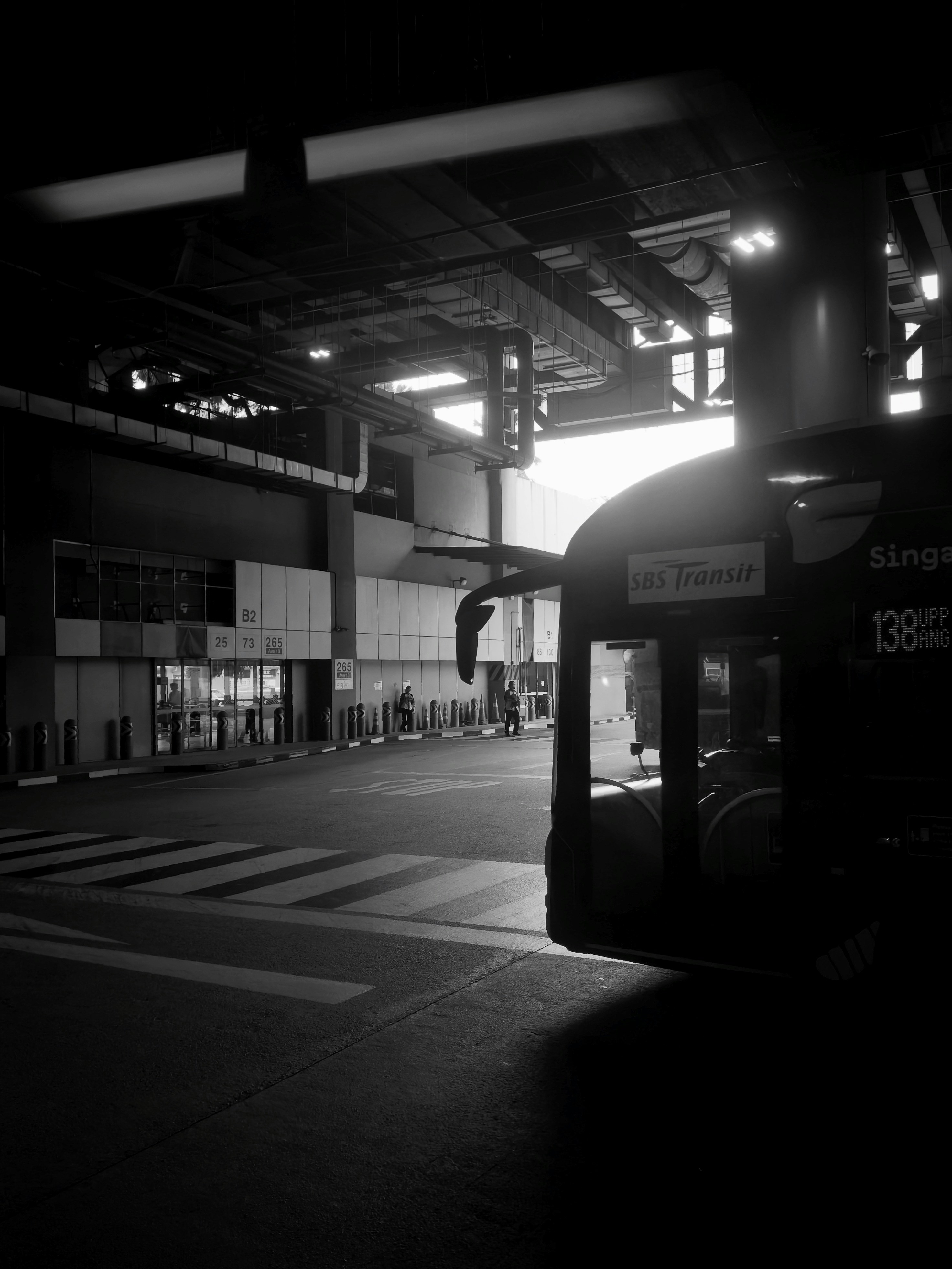 Dark, monochrome scene of a transit terminal with a bus on the right and a crosswalk leading into a bright exterior courtyard on the left; high-contrast lighting emphasizes architectural lines and depth.