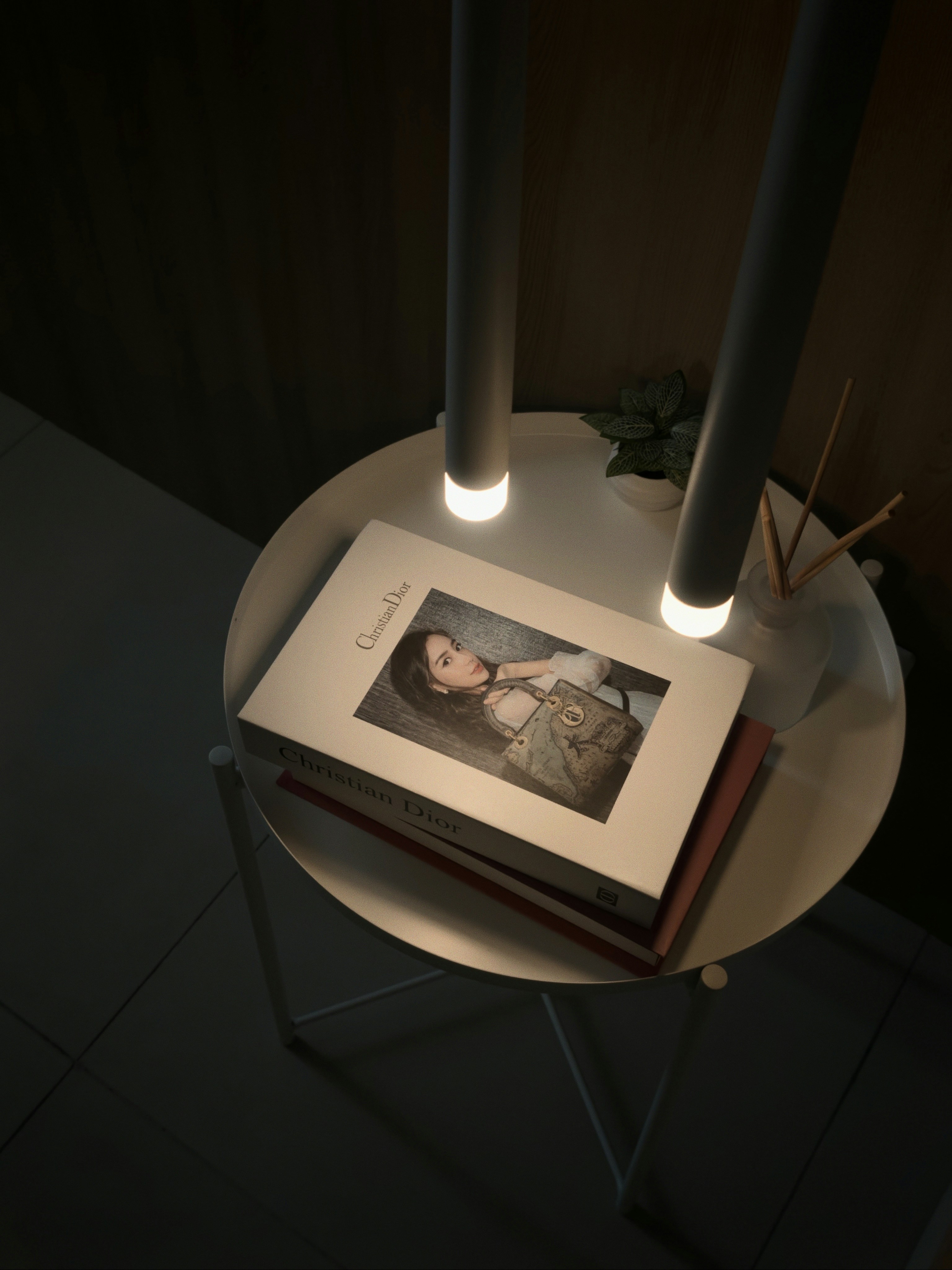 A small round side table with a stacked book and a boxed magazine sits under two vertical lamps, casting a warm glow on the page image; a few decorative items and a plant rest nearby.