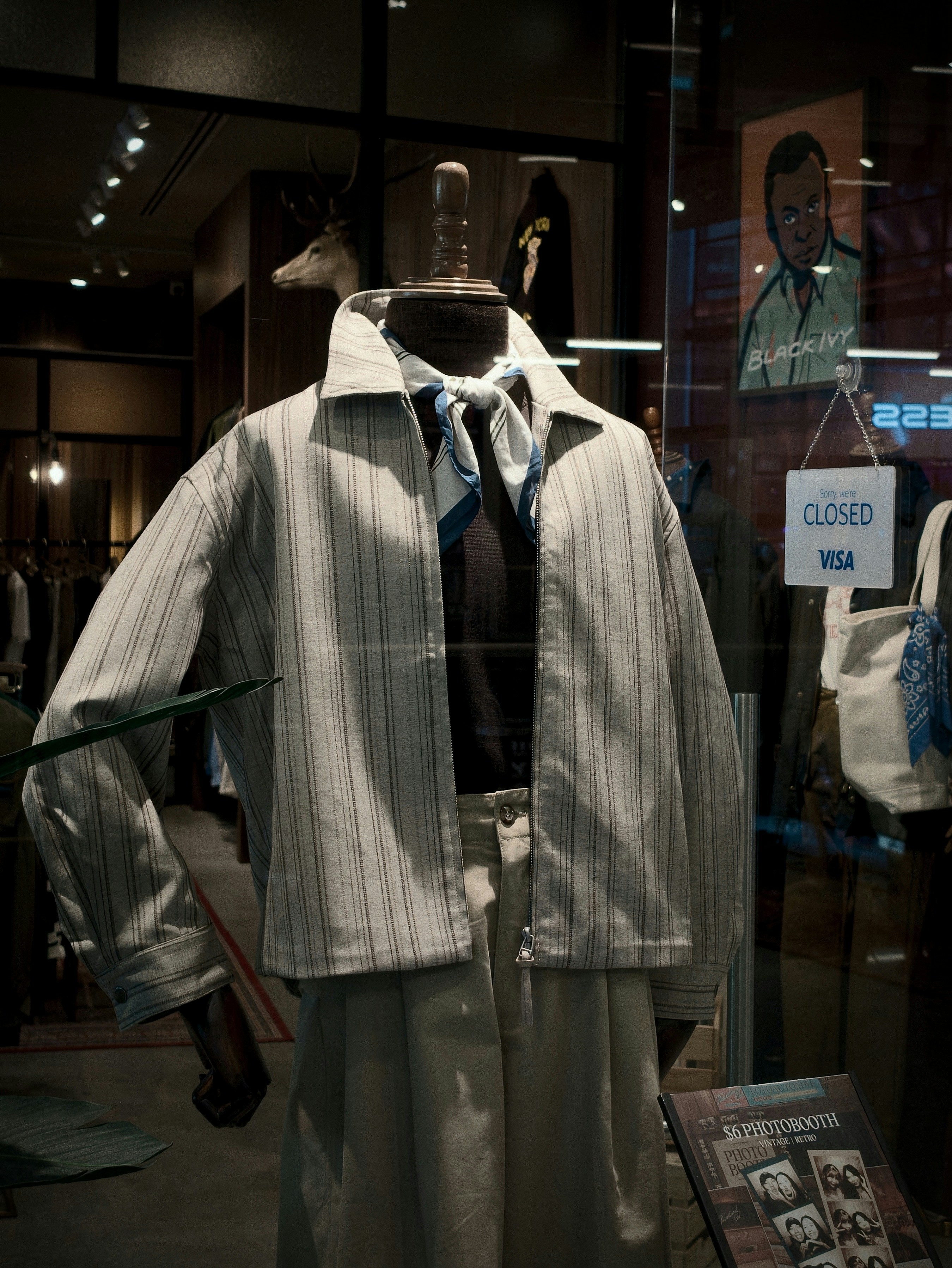 Glass-front display of a striped jacket and scarf on a mannequin inside a clothing store, with posters and signage visible in the background.