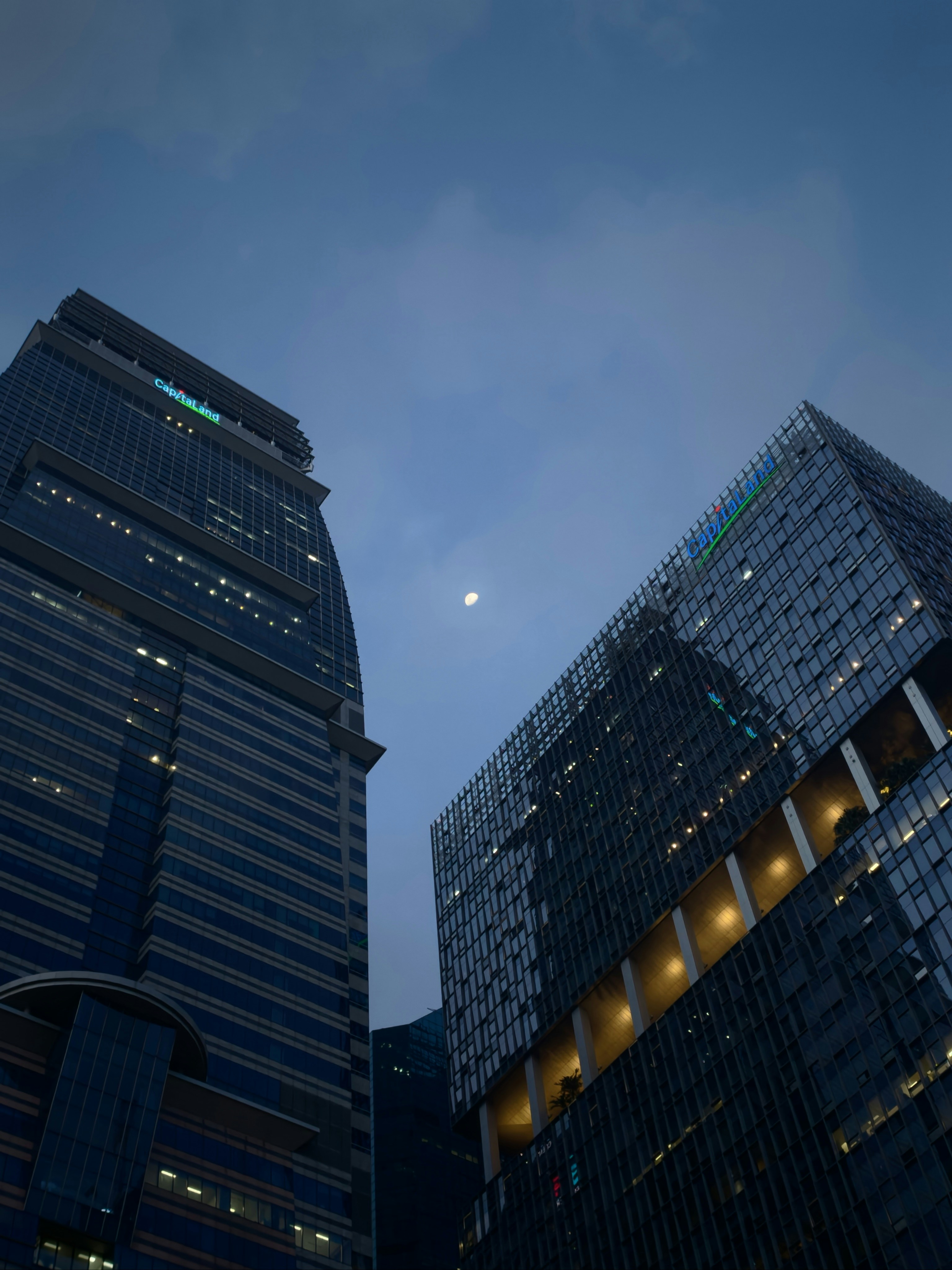 Two modern office towers reach toward a pale moon under a blue evening sky, contrasting glass façades reflecting subtle light. A vertical urban frame emphasizing geometry and scale.
