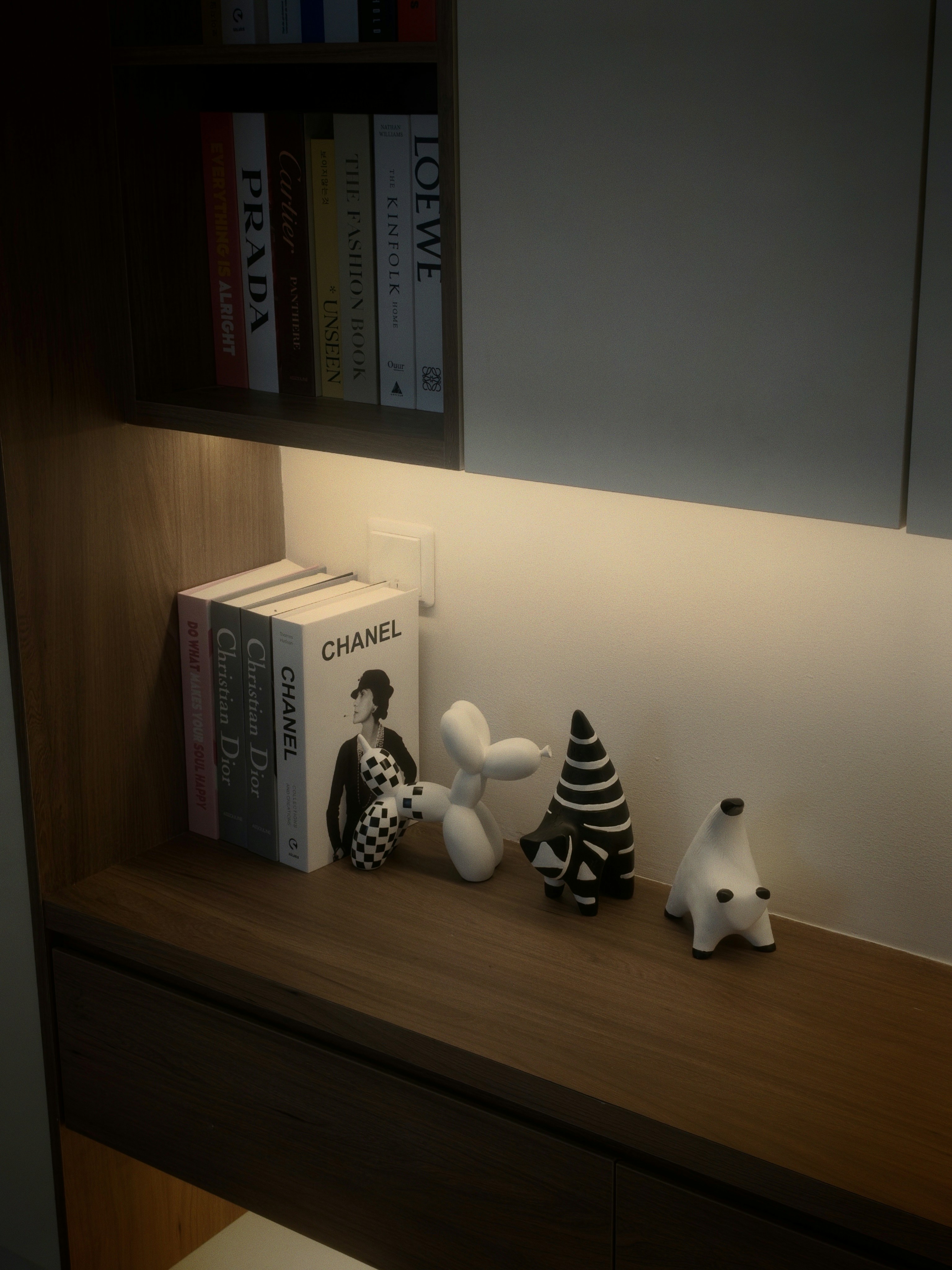 Arrangement of decorative ceramic figures and Chanel books on a warm-lit wooden shelf, with a soft backdrop and subtle shadows.