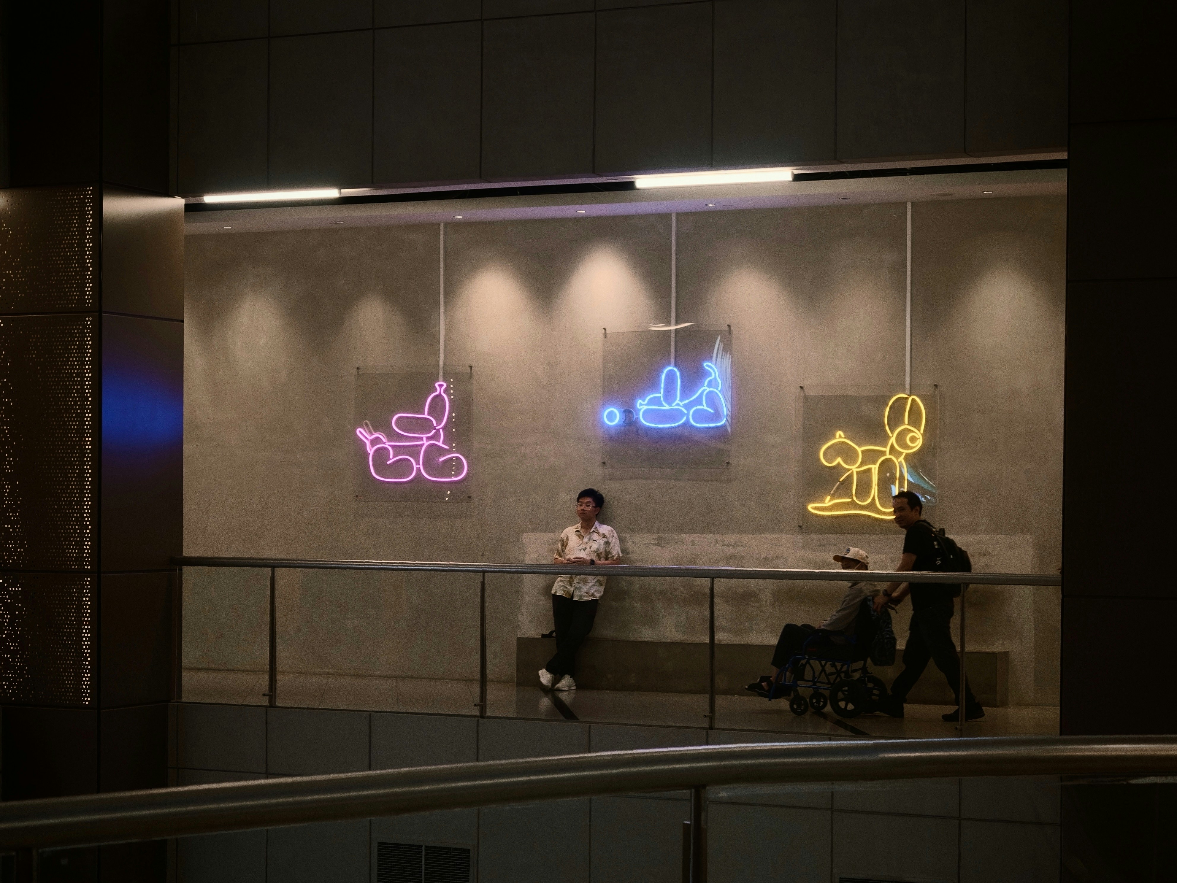 Neon outline art on a concrete wall behind a rail, with two people and a stroller along the walkway in a dim, modern indoor space.