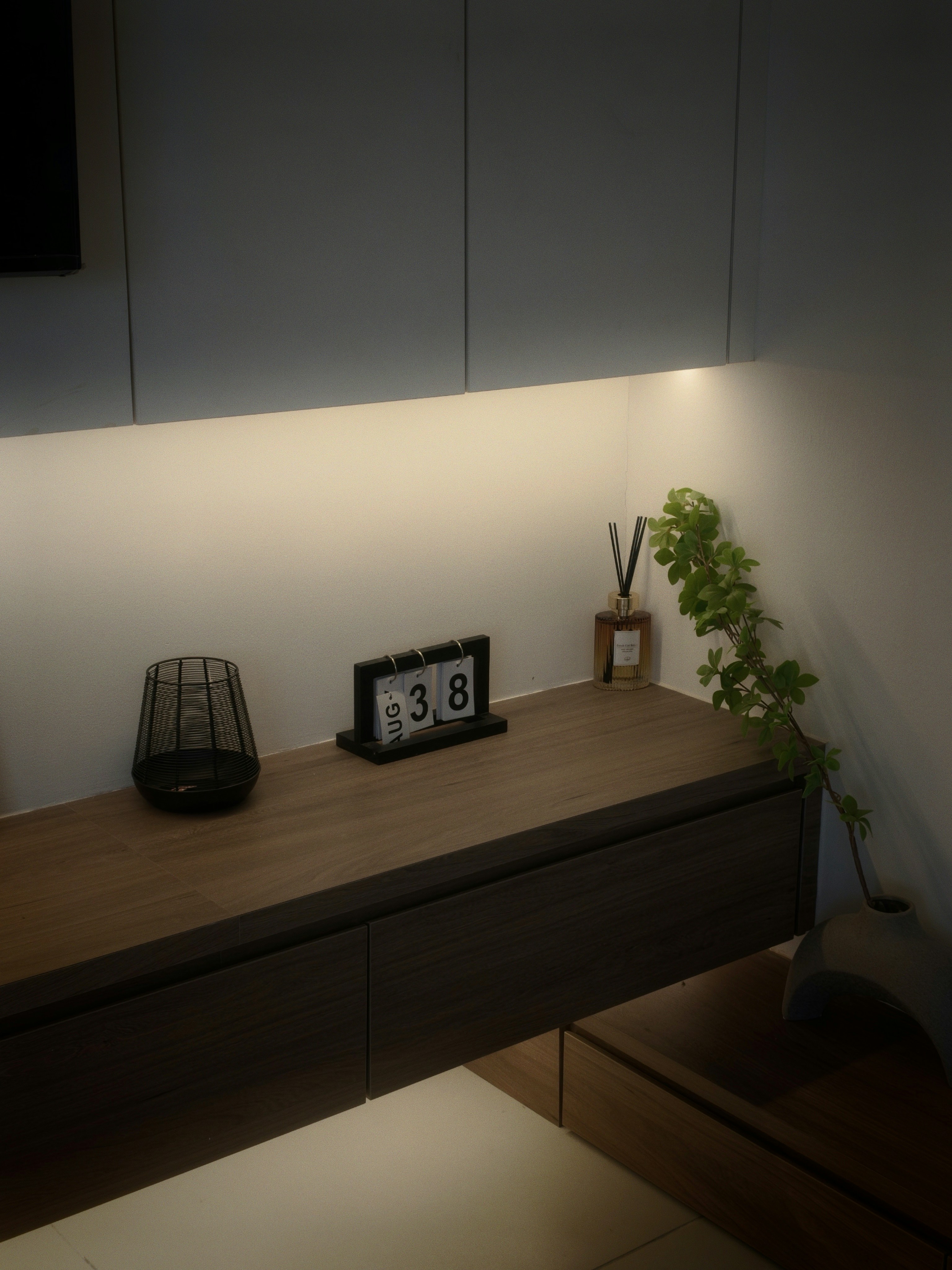 Warm, soft-lit kitchen console with dark wood cabinets, a small decorative clock, plant, and diffuser on a wooden countertop; minimal decor and ambient under-cabinet lighting.