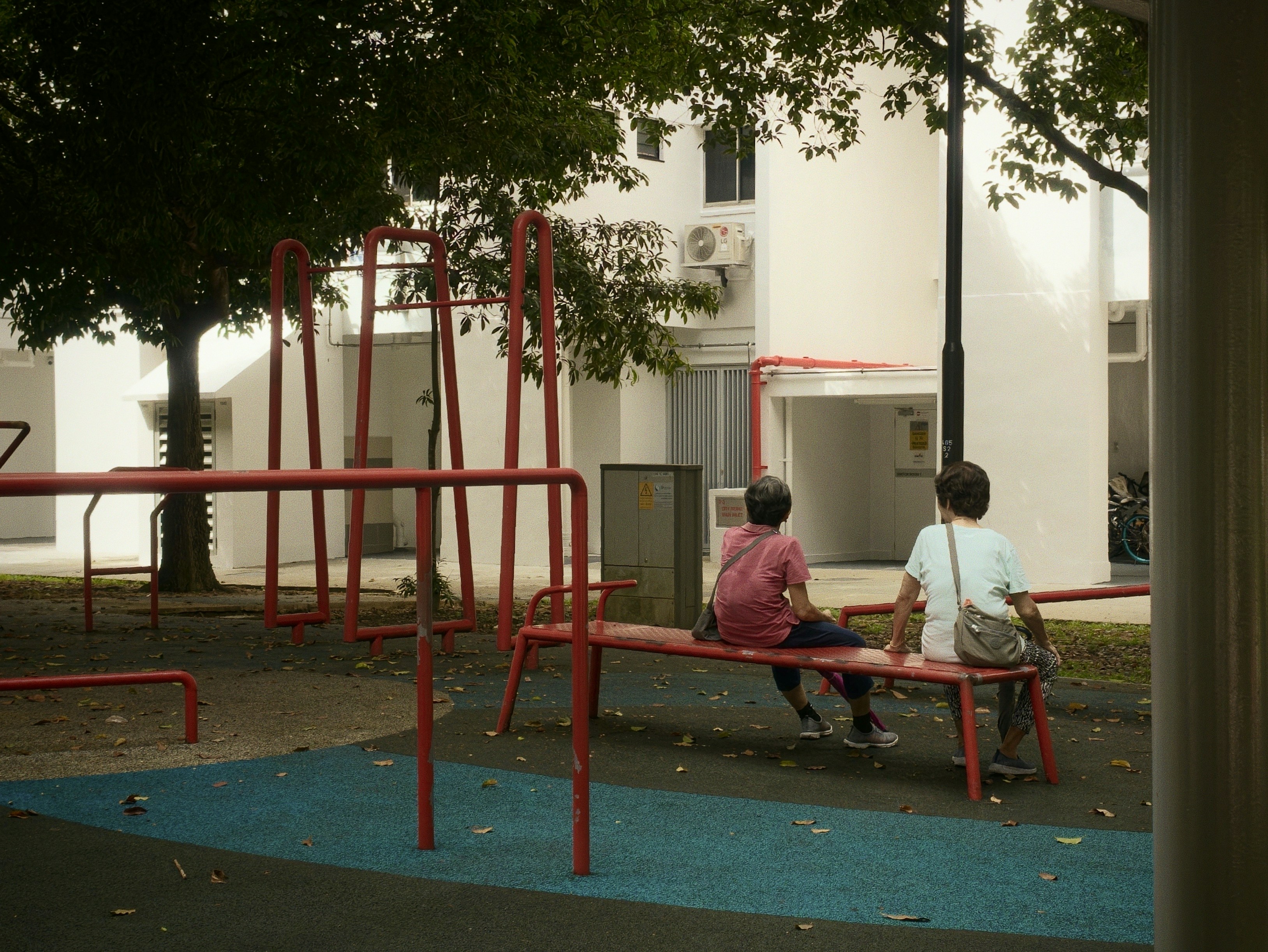 Two children sit on a red metal bench in a small urban playground; a blue rubberized strip and red structures frame the scene, while a building and trees provide the backdrop.