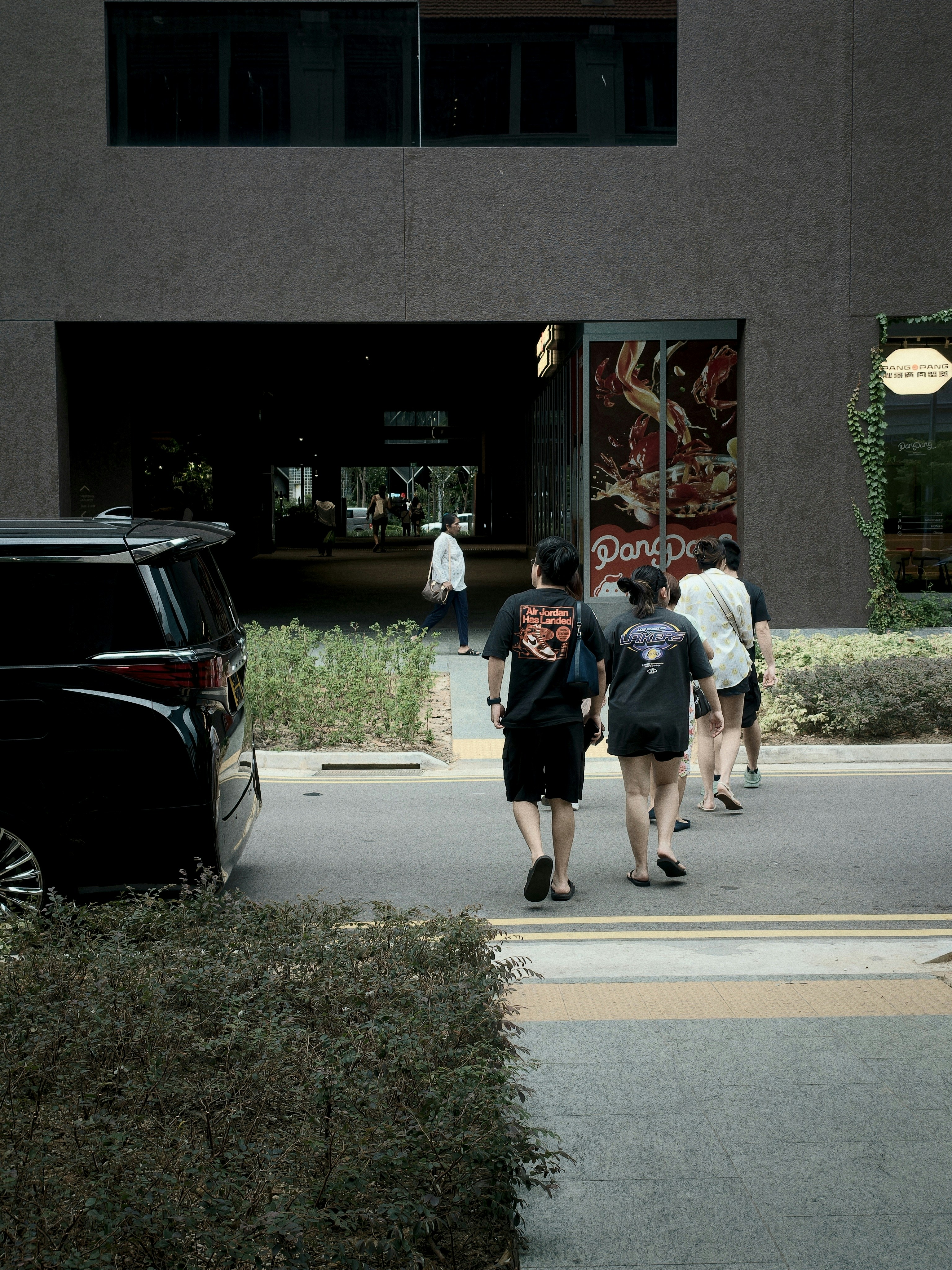 Young pedestrians walk toward a dimly lit building entrance across a street, with a parked vehicle on the left and a poster on the right; the scene captures a candid urban moment.