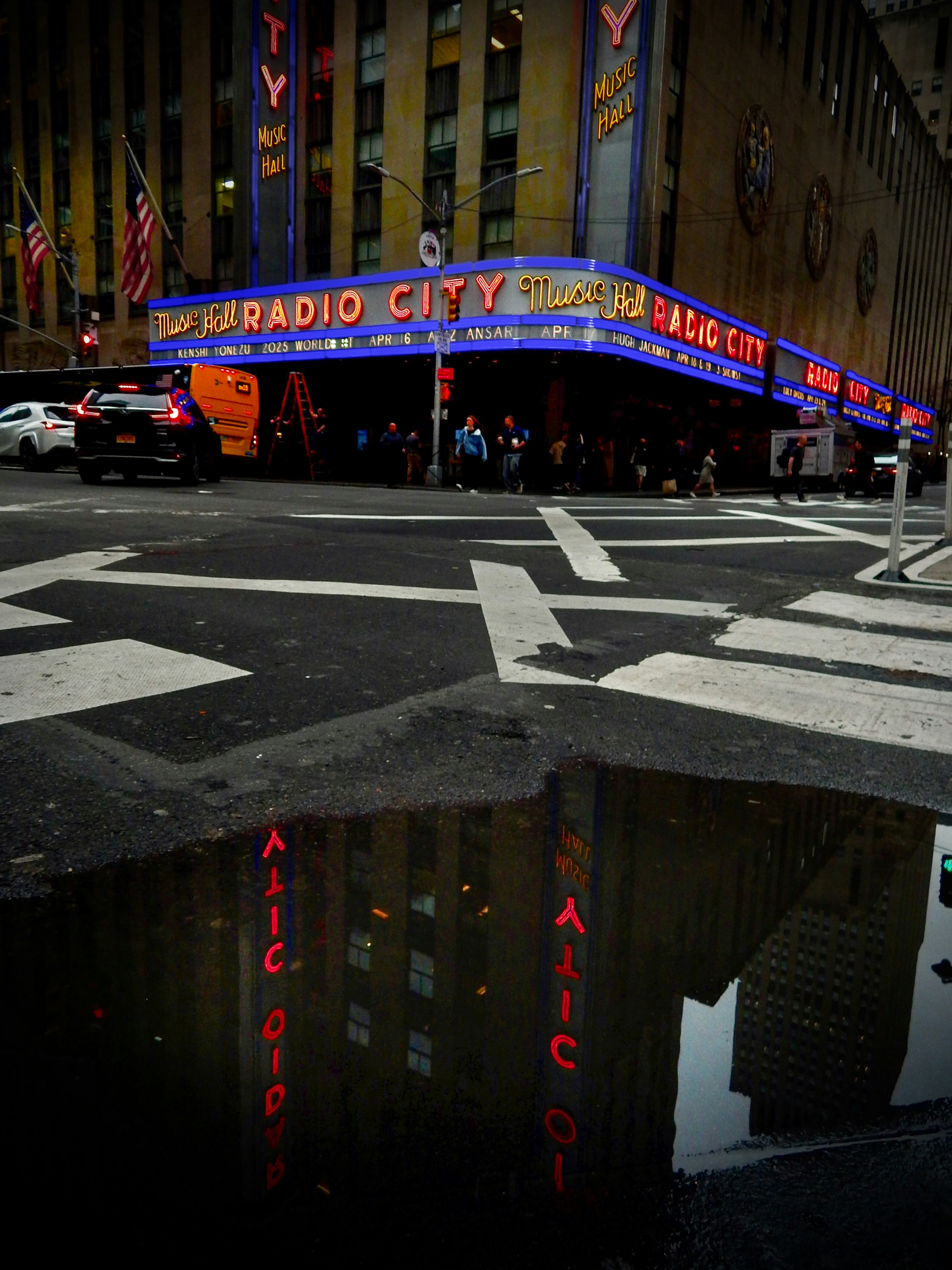 Neon-lit Radio City Music Hall facade at dusk with street crossing, reflected sign in a puddle that doubles the vertical composition.
