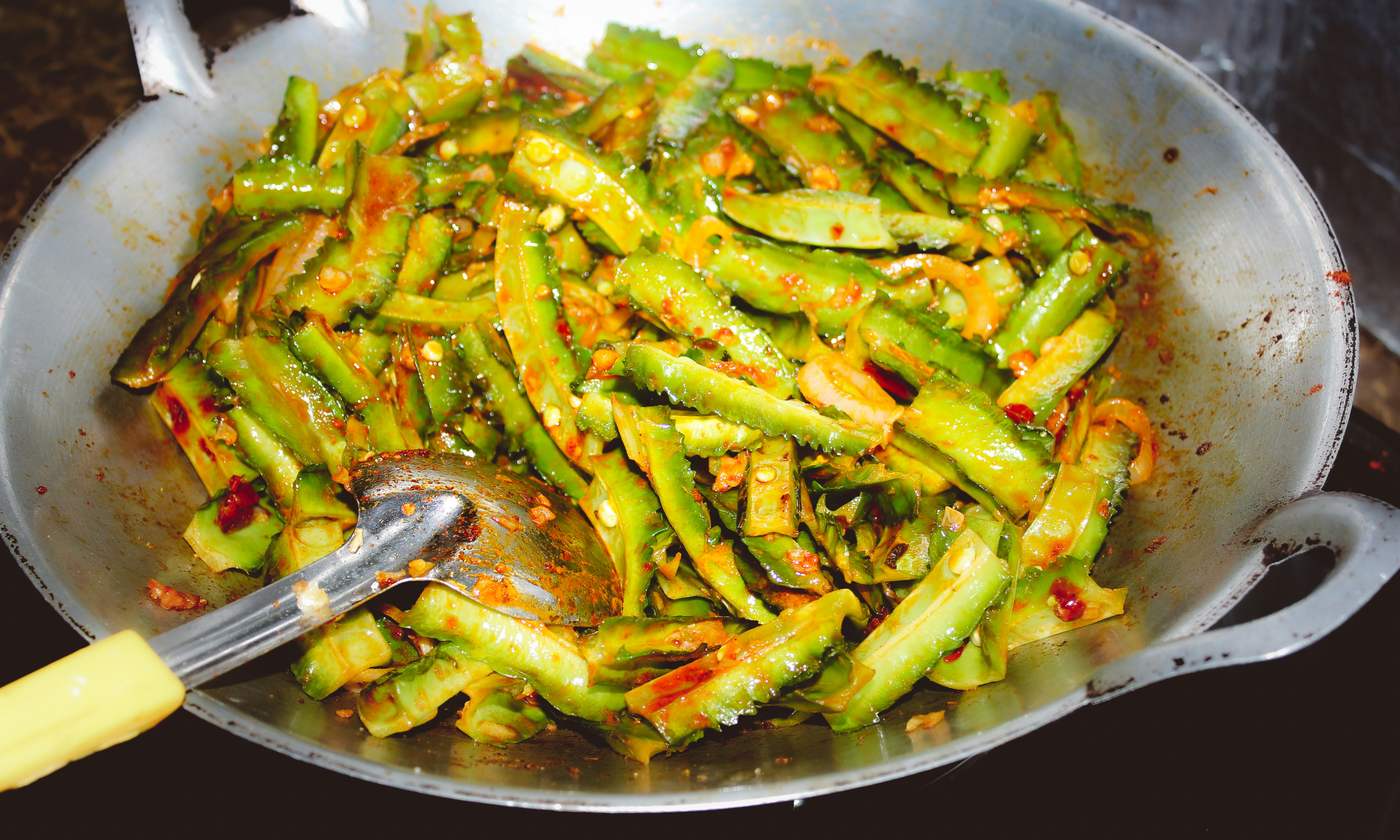 Bright green vegetable medley sizzling in a wide pan with red chili accents; a spatula rests in the wok, suggesting active cooking. A culinary scene focused on stir-fry ingredients and technique.
