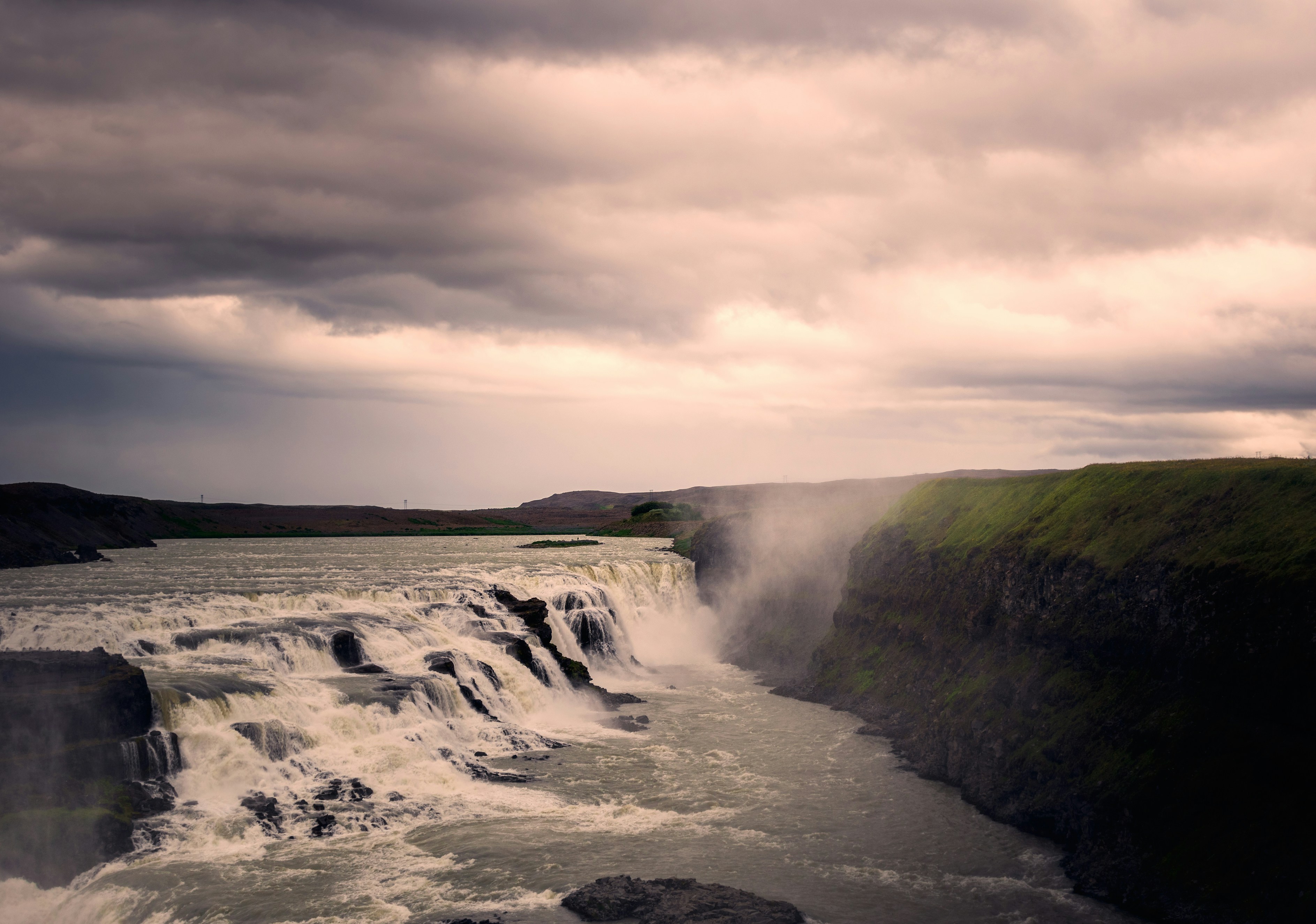 Misty waterfall spills over a dark cliff into a churning river under a moody sky; distant horizon and grassy ledge add depth.