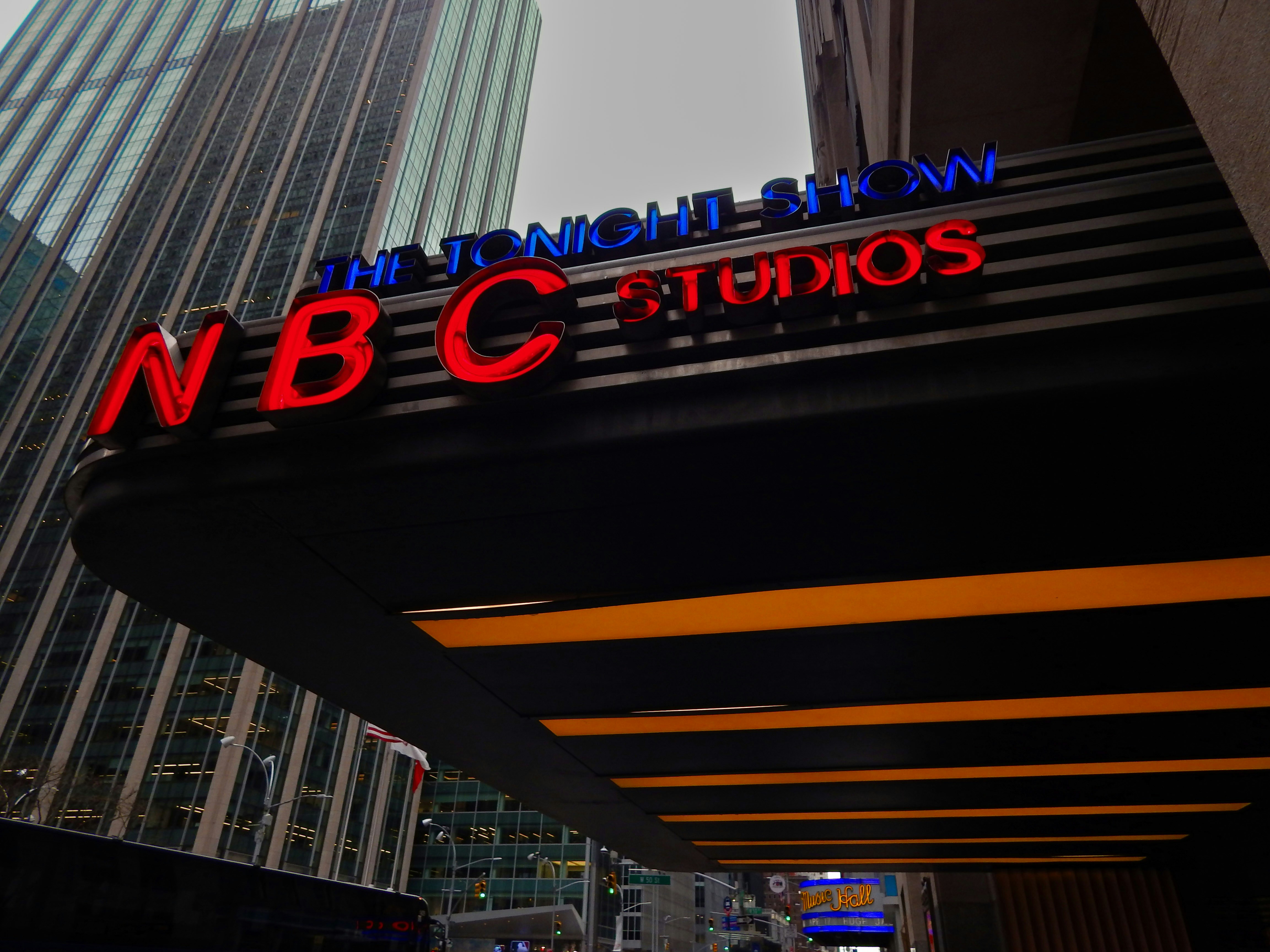 Neon NBC Studios sign over a city street with a dark canopy and vertical glass towers in the backdrop.