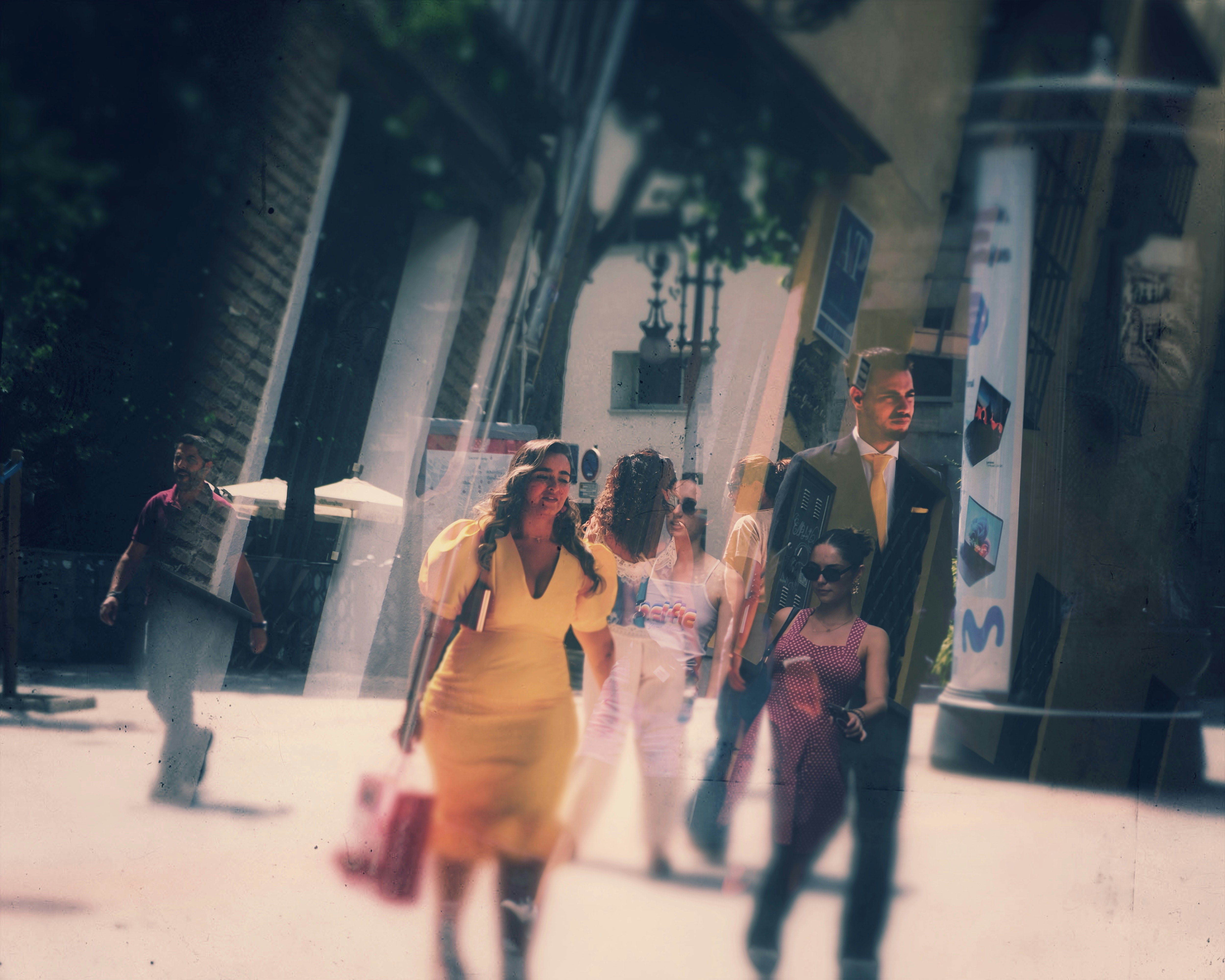 A blurred street scene shows well-dressed pedestrians walking toward a sunlit avenue, with a bright yellow-clad woman near the center drawing attention. A logo/branding element appears on a vertical signpost to the right, while overall warmth and shallow focus give a dreamy mood.