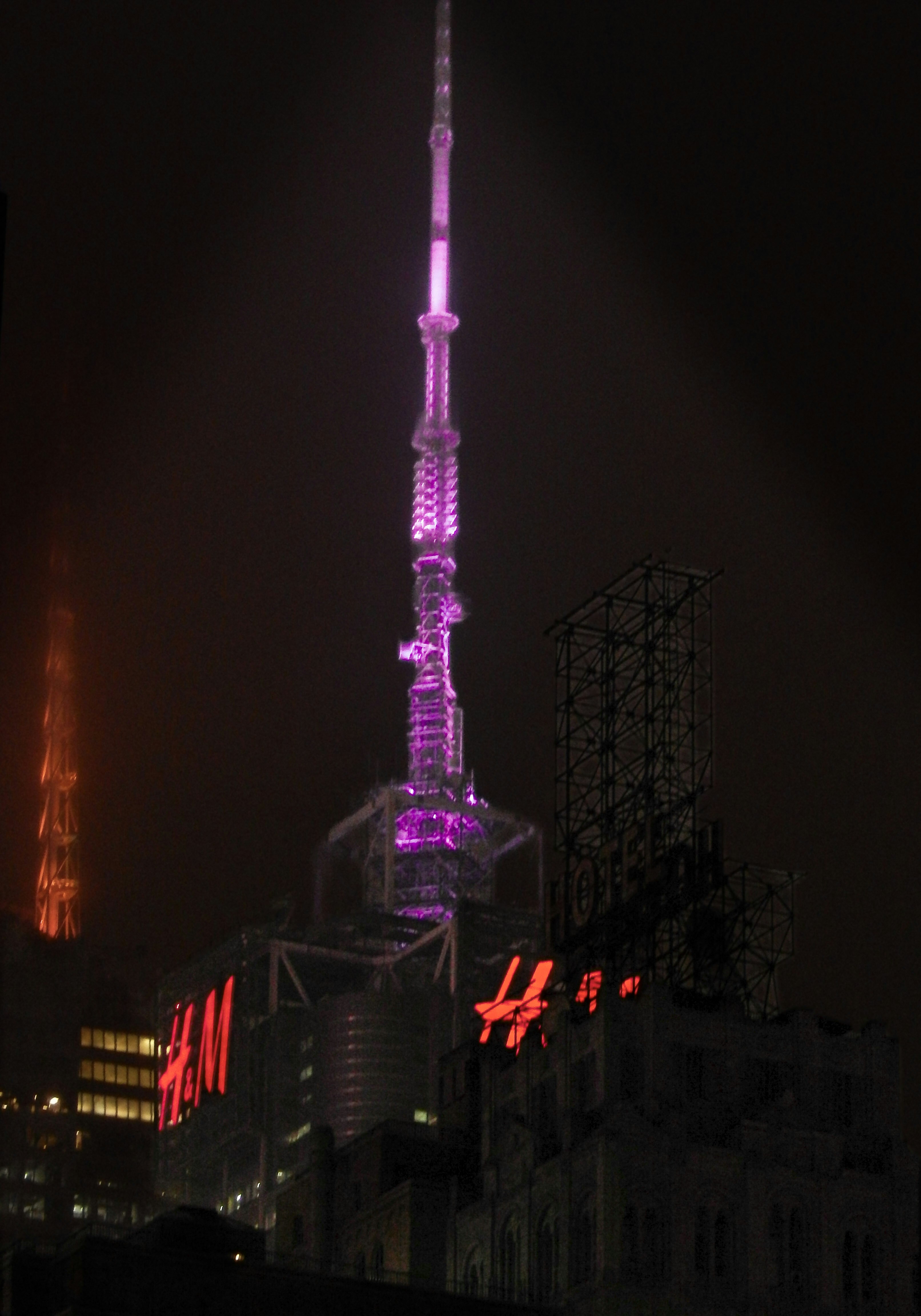 Night scene of a tall, illuminated communications tower with magenta lighting atop a city building; signs glow red while surrounding structures fade into darkness.