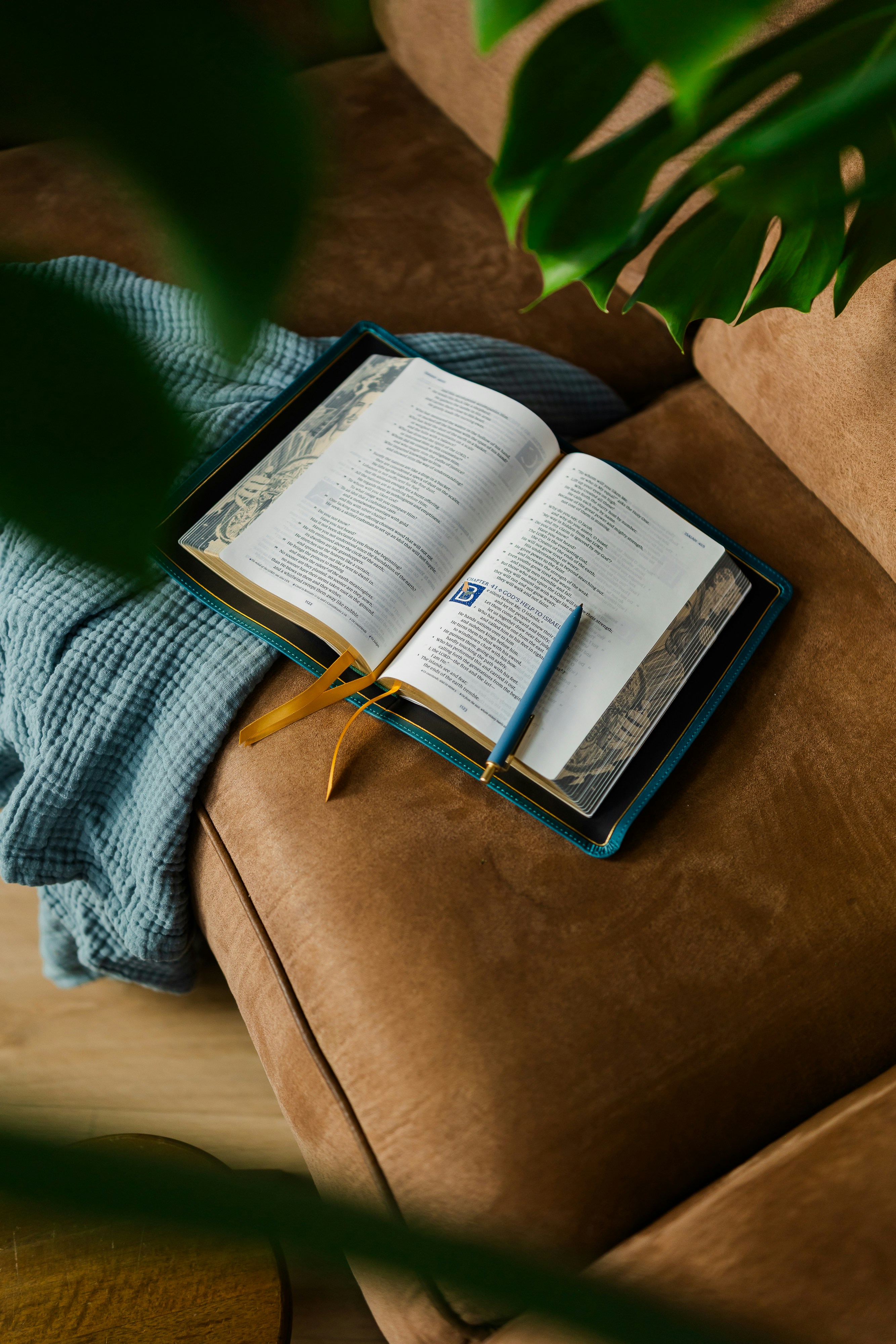Open book with a blue pencil on a brown couch, partially covered by a textured teal blanket; soft natural light and plant leaves frame the scene. This is a lifestyle photograph featuring a printed book and stationery.