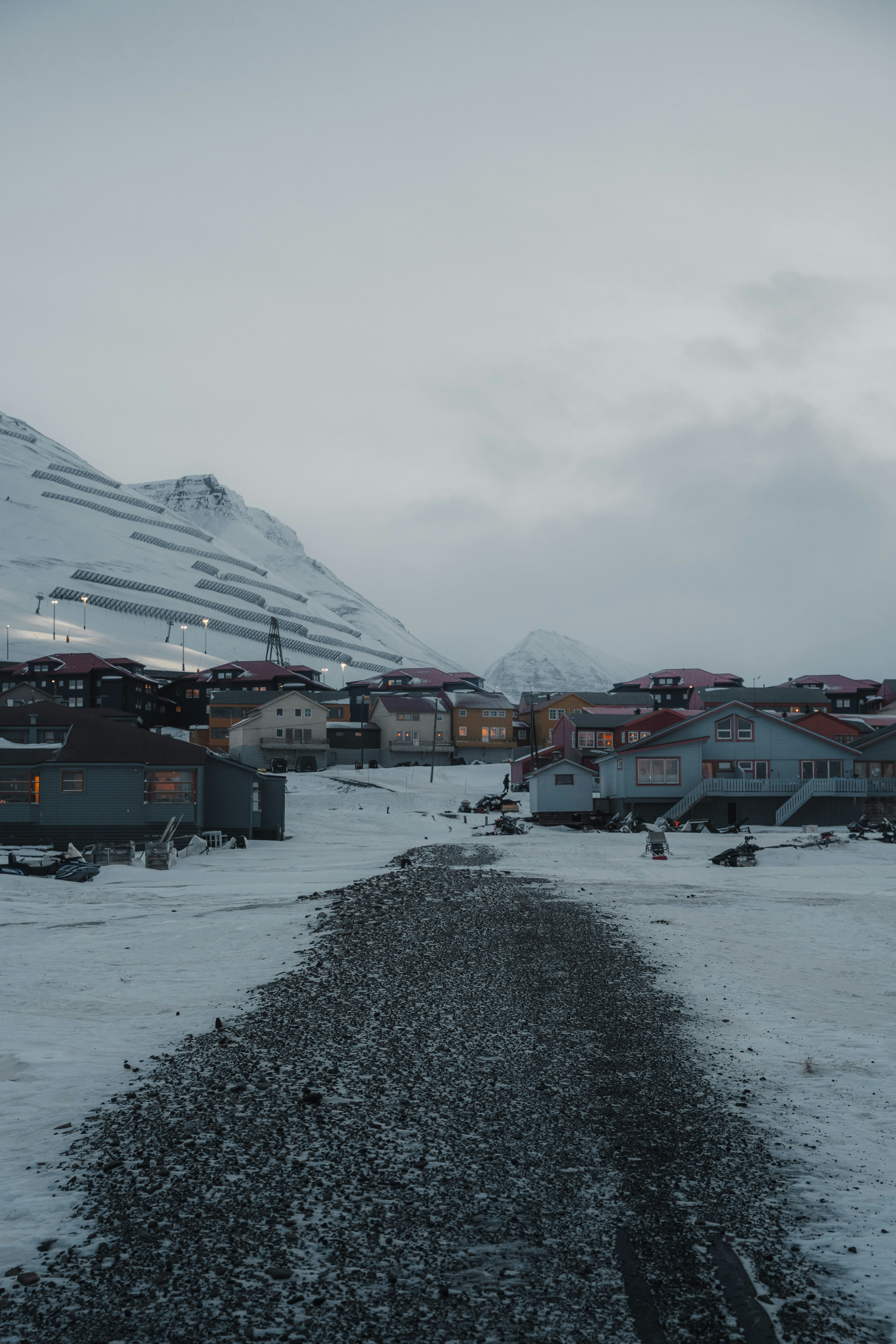 Snow-covered village with colorful houses along a rocky path toward distant mountains under a pale sky. The scene emphasizes winter lighting and quiet, residential detail in a remote Arctic setting.