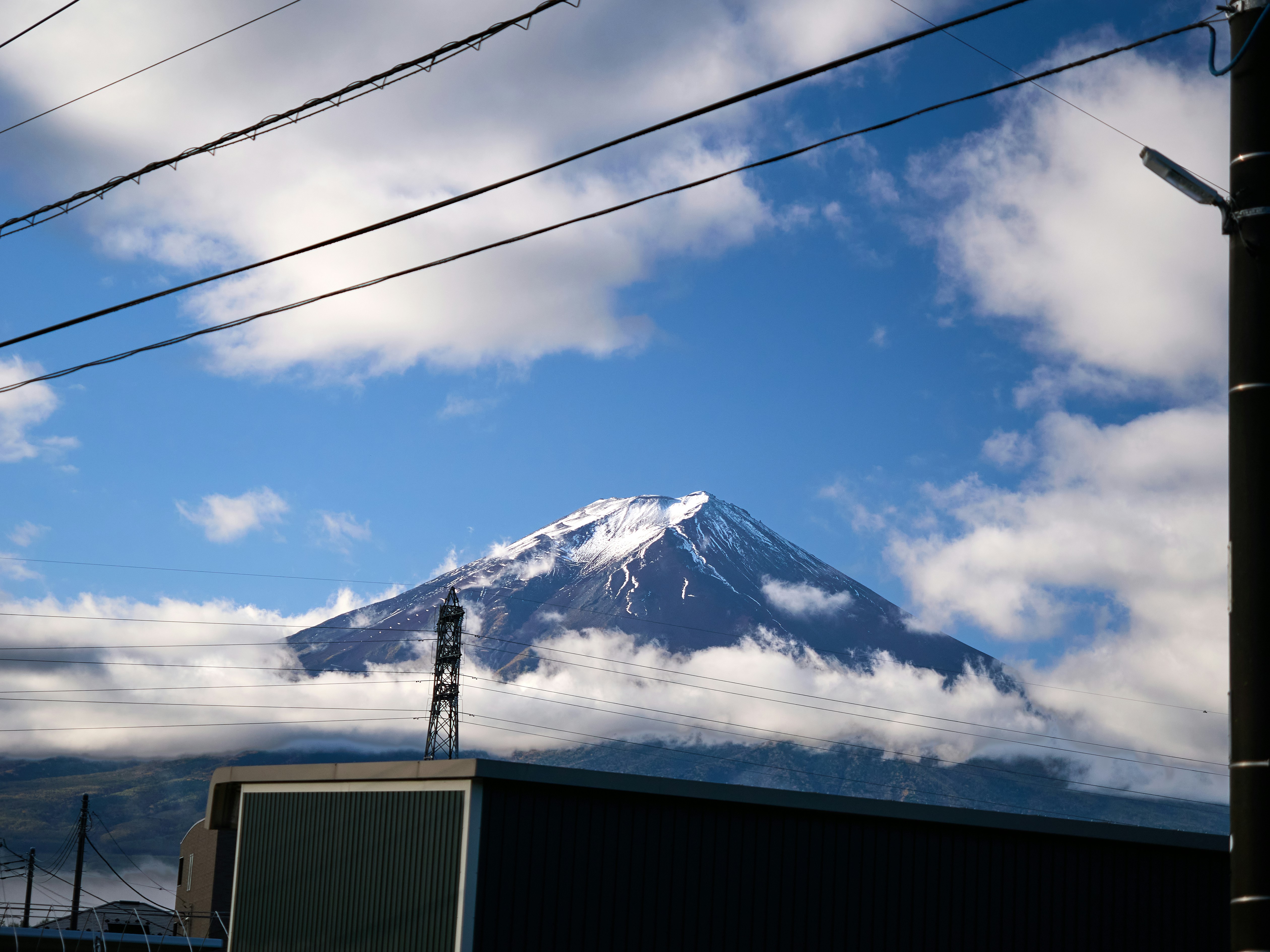Snowy mountain peak peeks above a low-lying cloud line, with power lines and a dark industrial building in the foreground.