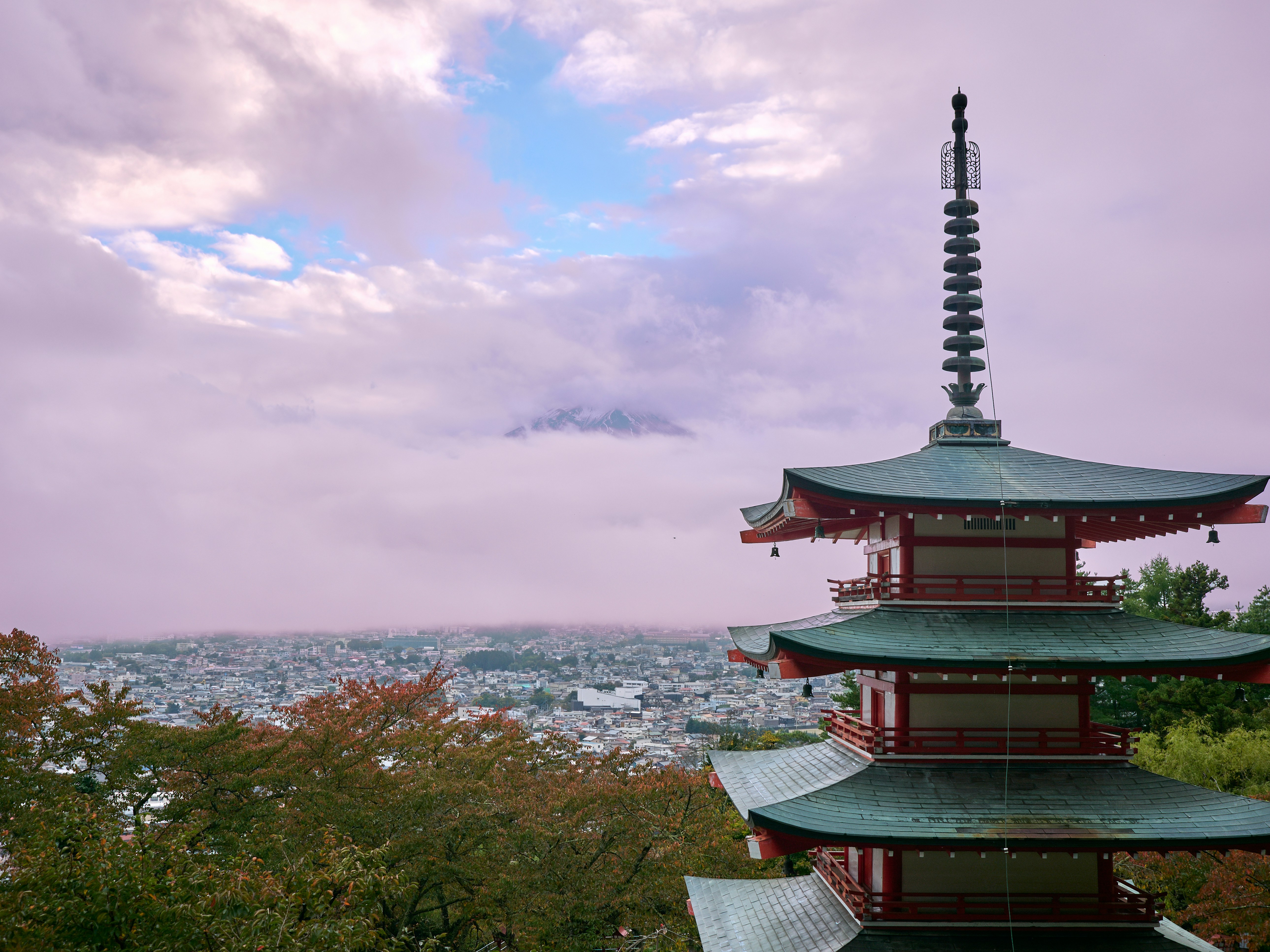 Three-tier pagoda perched on a hillside overlooking a distant city, with autumn foliage and a cloudy sky.