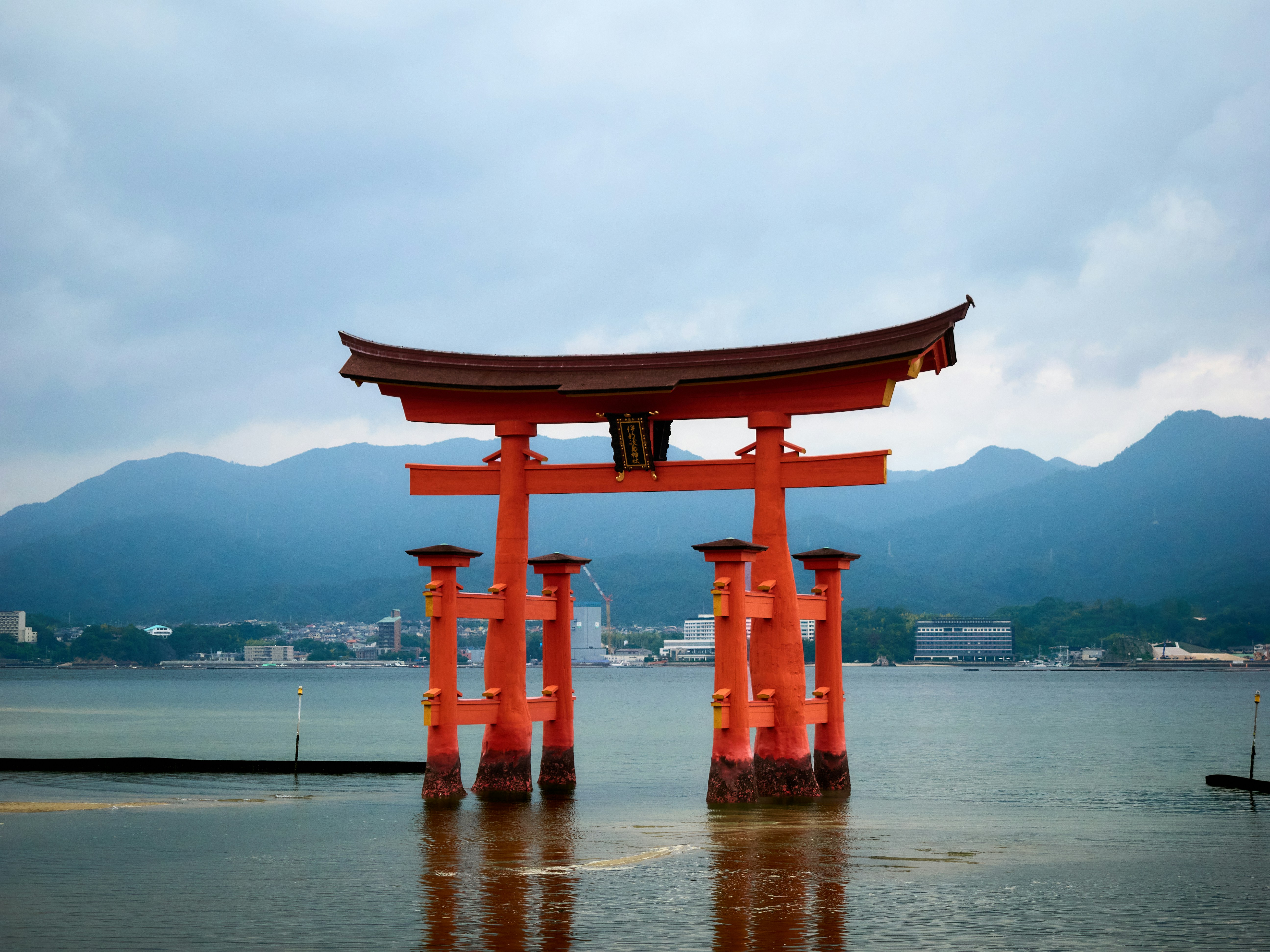 Bright orange torii rise from calm shallow water with distant mountains, framing a tranquil coastal scene.