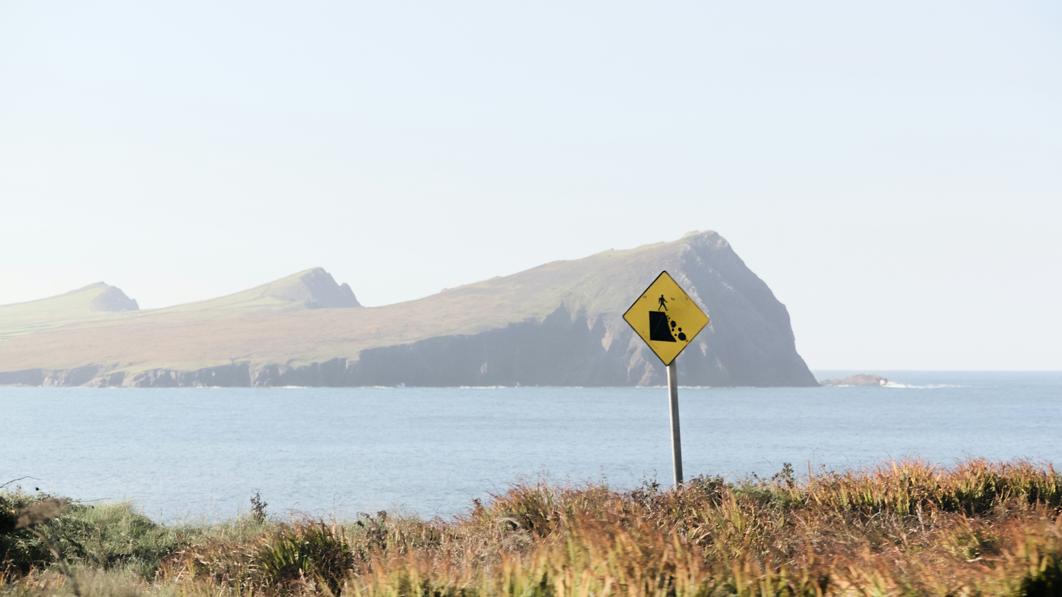 Coastal landscape with a yellow warning sign by the sea, grasses in the foreground and distant cliffs under a clear sky.