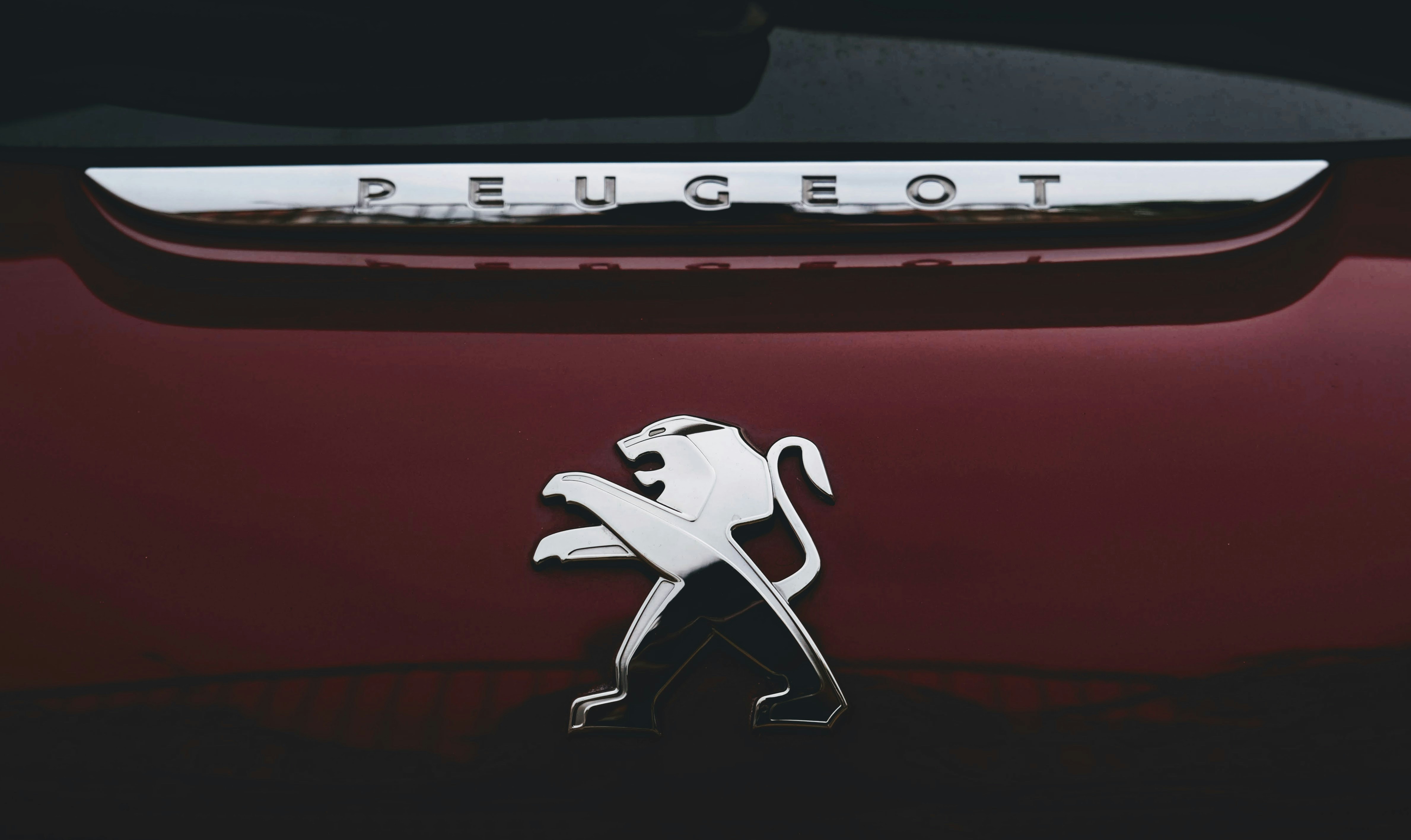 Close-up of a Peugeot badge on a deep red car with a chrome strip above; glossy paint and soft reflections emphasize the emblem as the focal point.