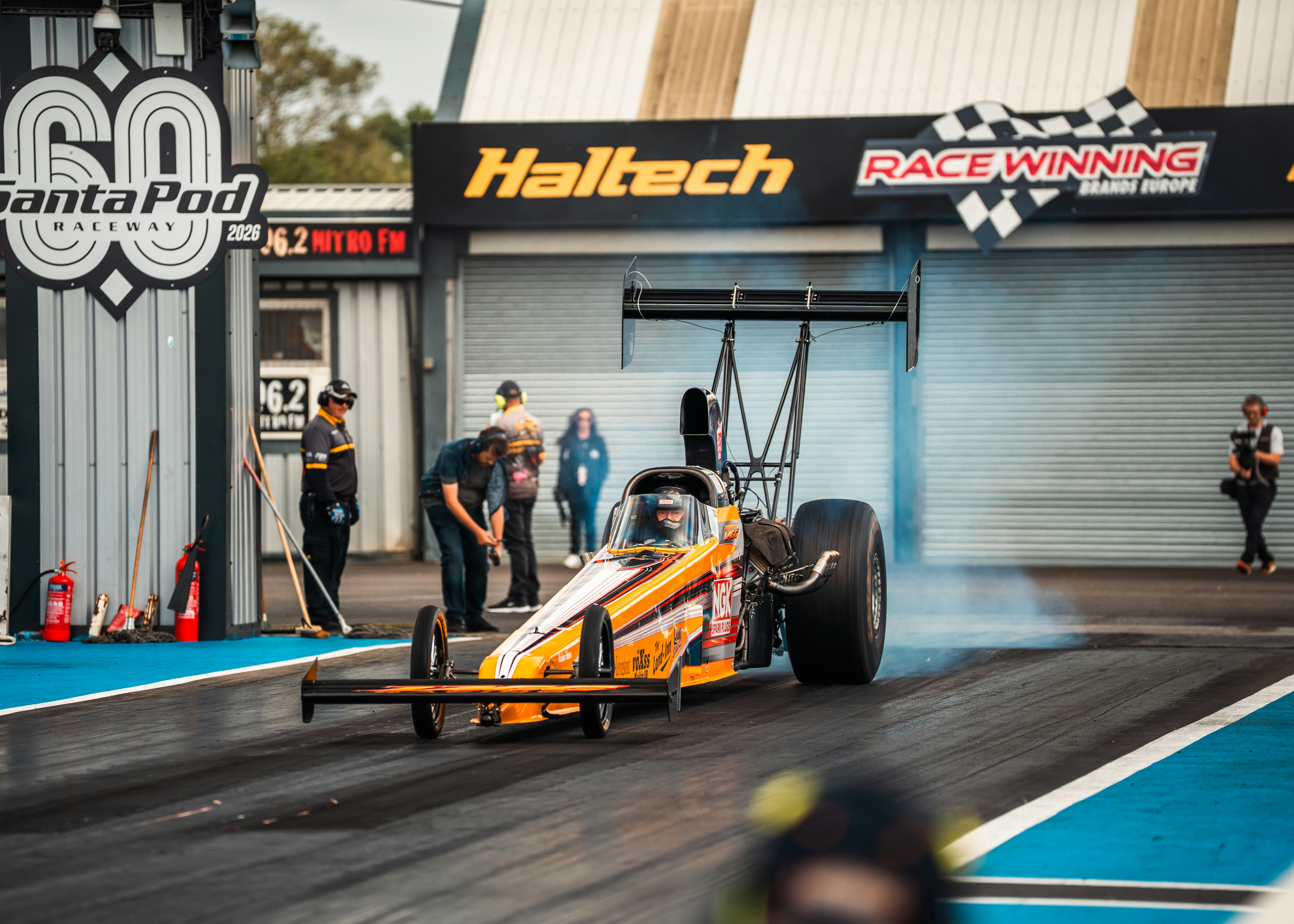 Orange nitro dragster at the starting line, smoke trailing as it powers down a raceway with team members and banners in the background.