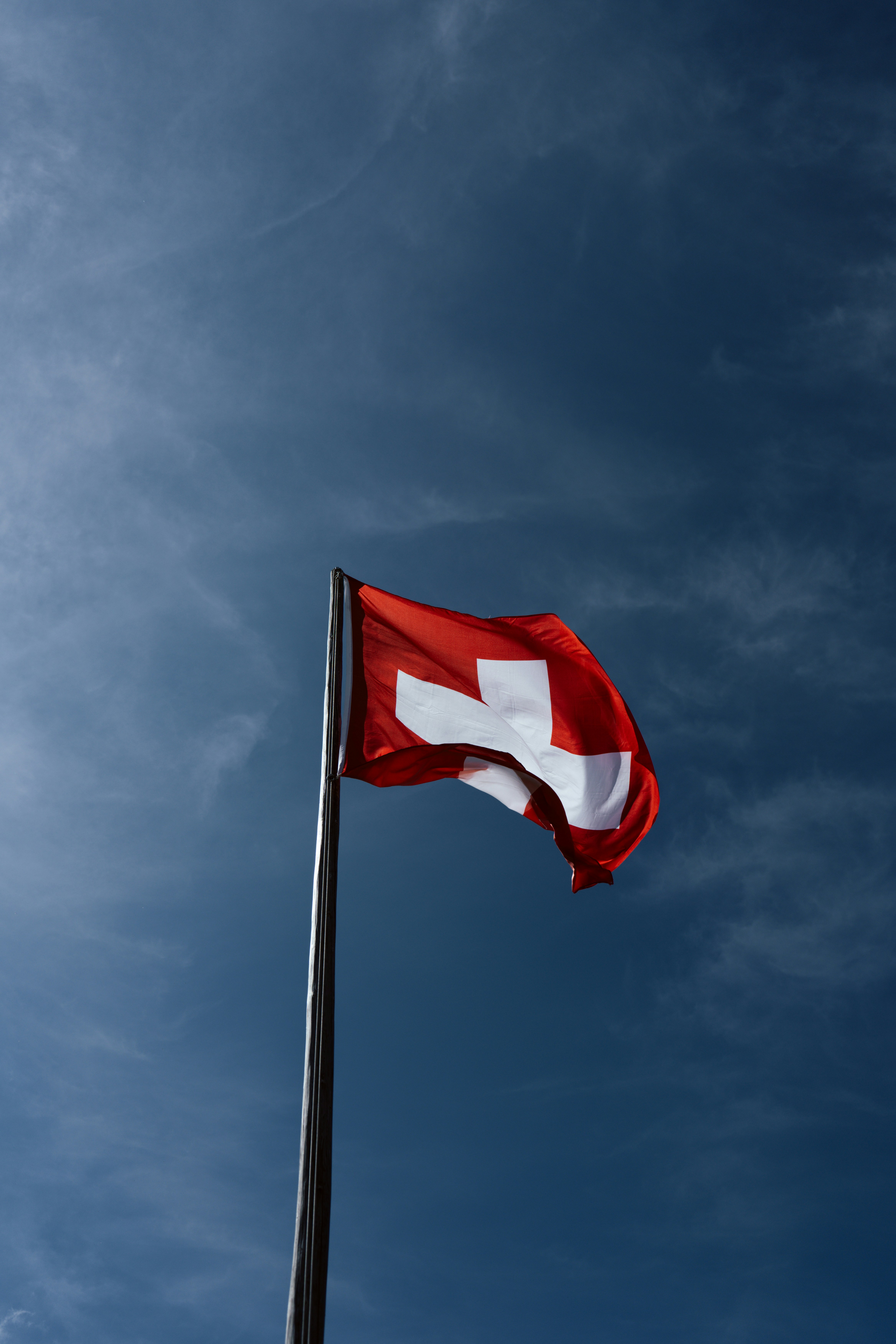 Swiss flag waves on a tall flagpole against a deep blue sky, photographed from below to emphasize height and contrast. The image focuses on the flag as the primary subject; suitable for architectural or photographic composition highlights.