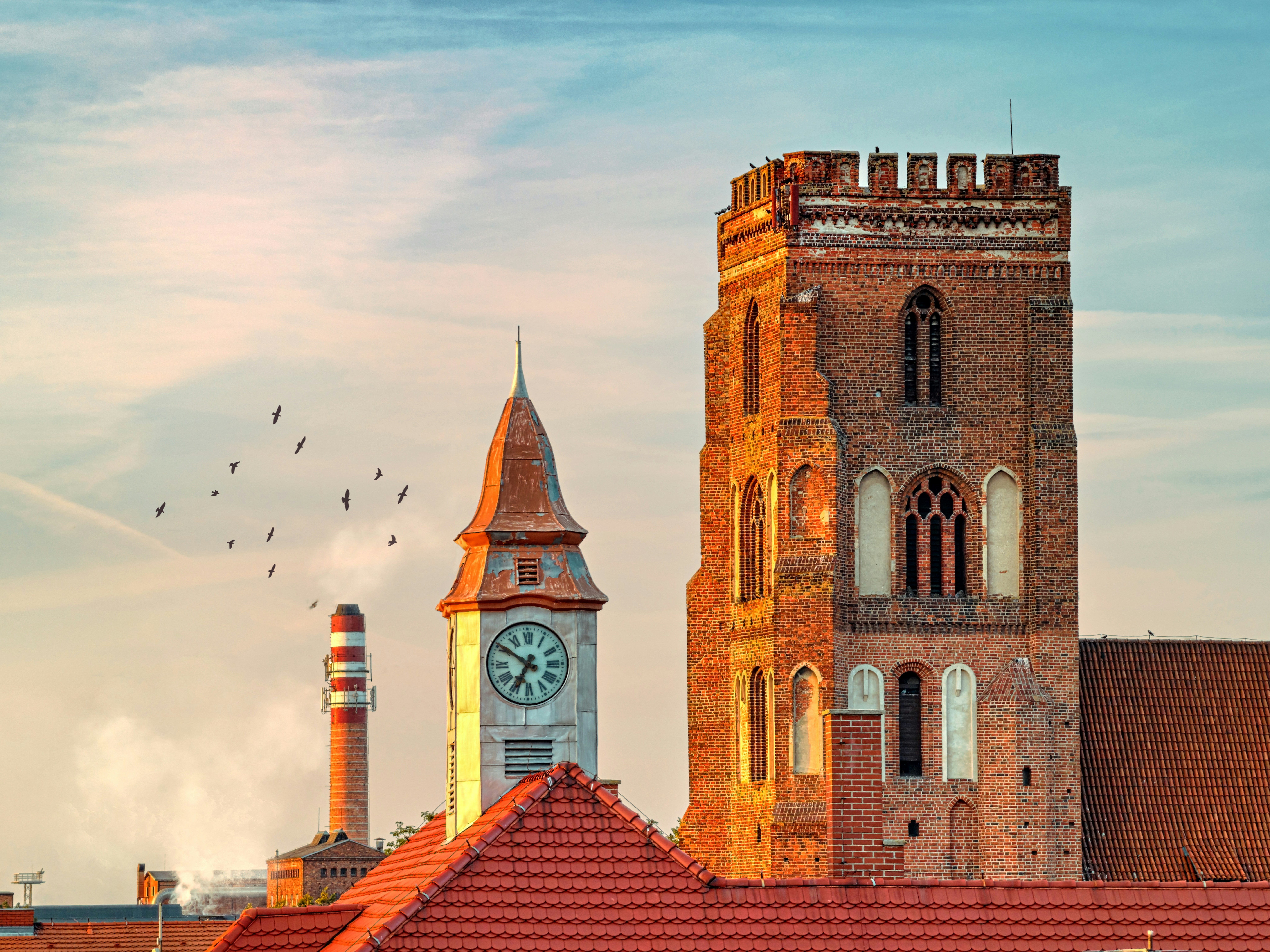 Brick church towers rise above red tiled rooftops as the clock tower catches the warm evening light; distant factory chimney and flock of birds add atmosphere to the skyline.