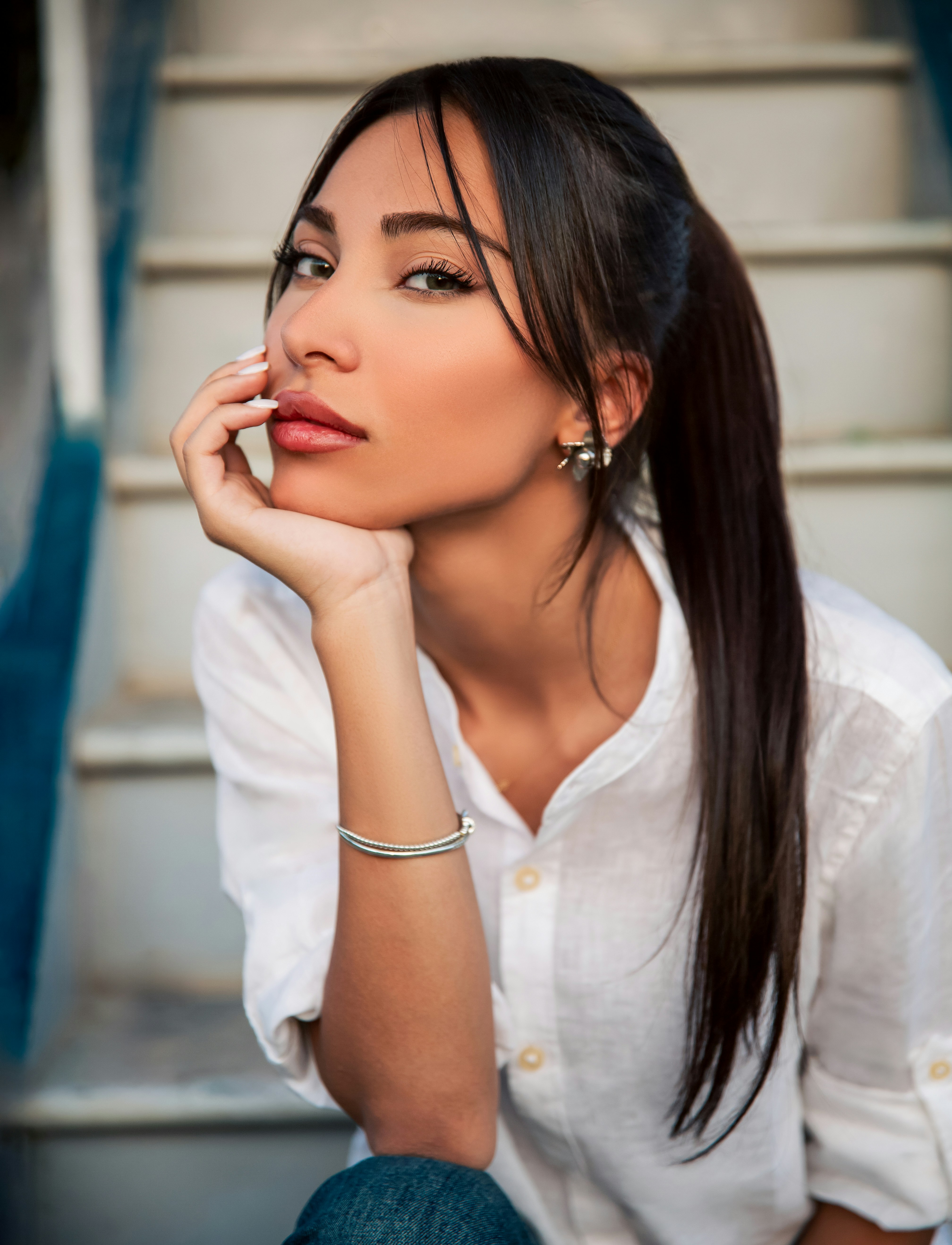 Soft portrait of a woman sitting on stairs in a white shirt, resting her chin on her hand; natural light highlights her features. This is a photographic portrait, not a painting or illustration.