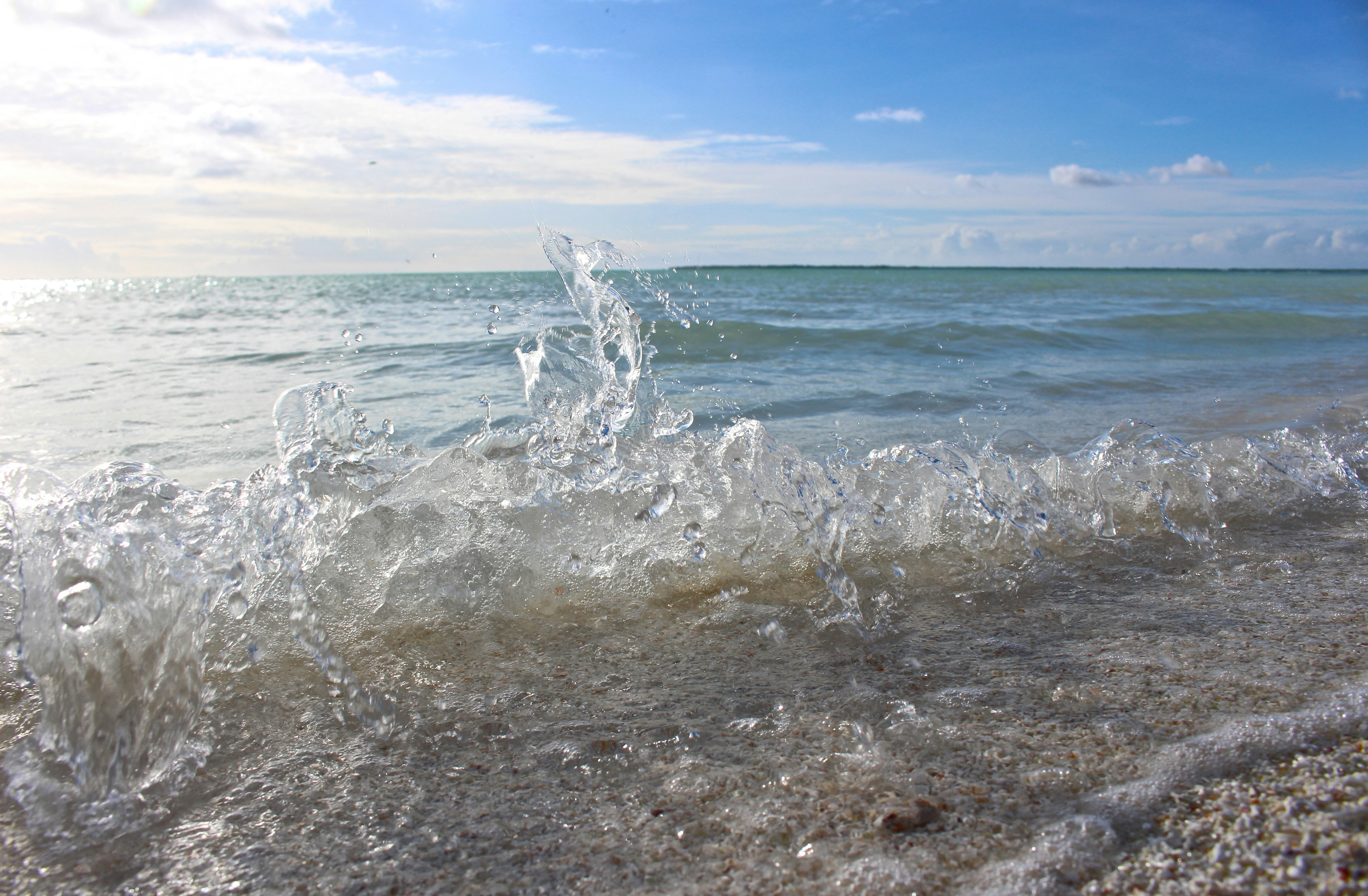 Close-up of a wave breaking over pebbles along a sunlit sea shore; the horizon and blue sky form the backdrop for the foamy spray and shallow-water detail.