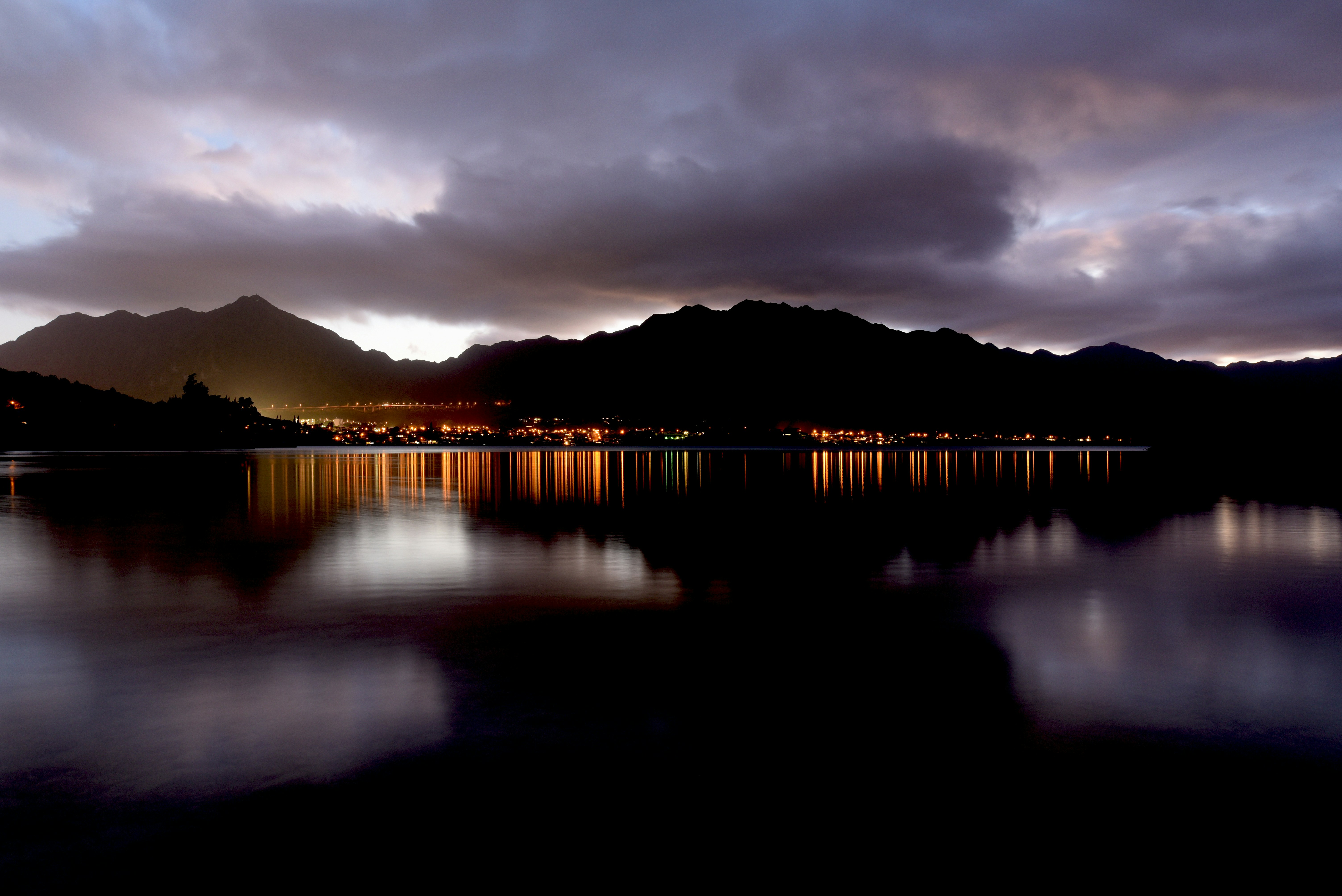 Night lakeside scene with distant mountain silhouettes and a row of illuminated buildings reflecting in calm water.