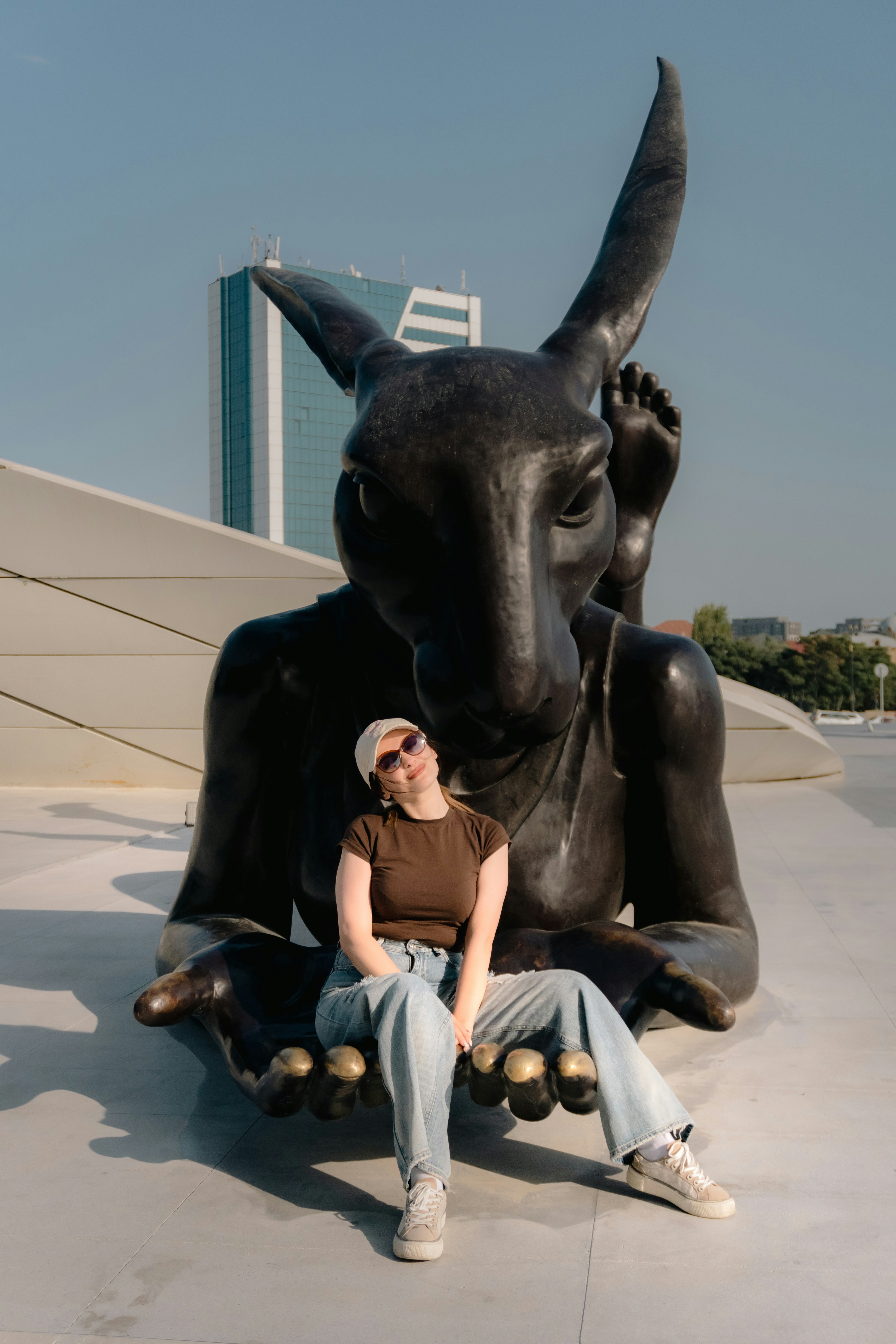 Girl in casual wear sits within the statue’s open hands, with a futuristic tower in the background