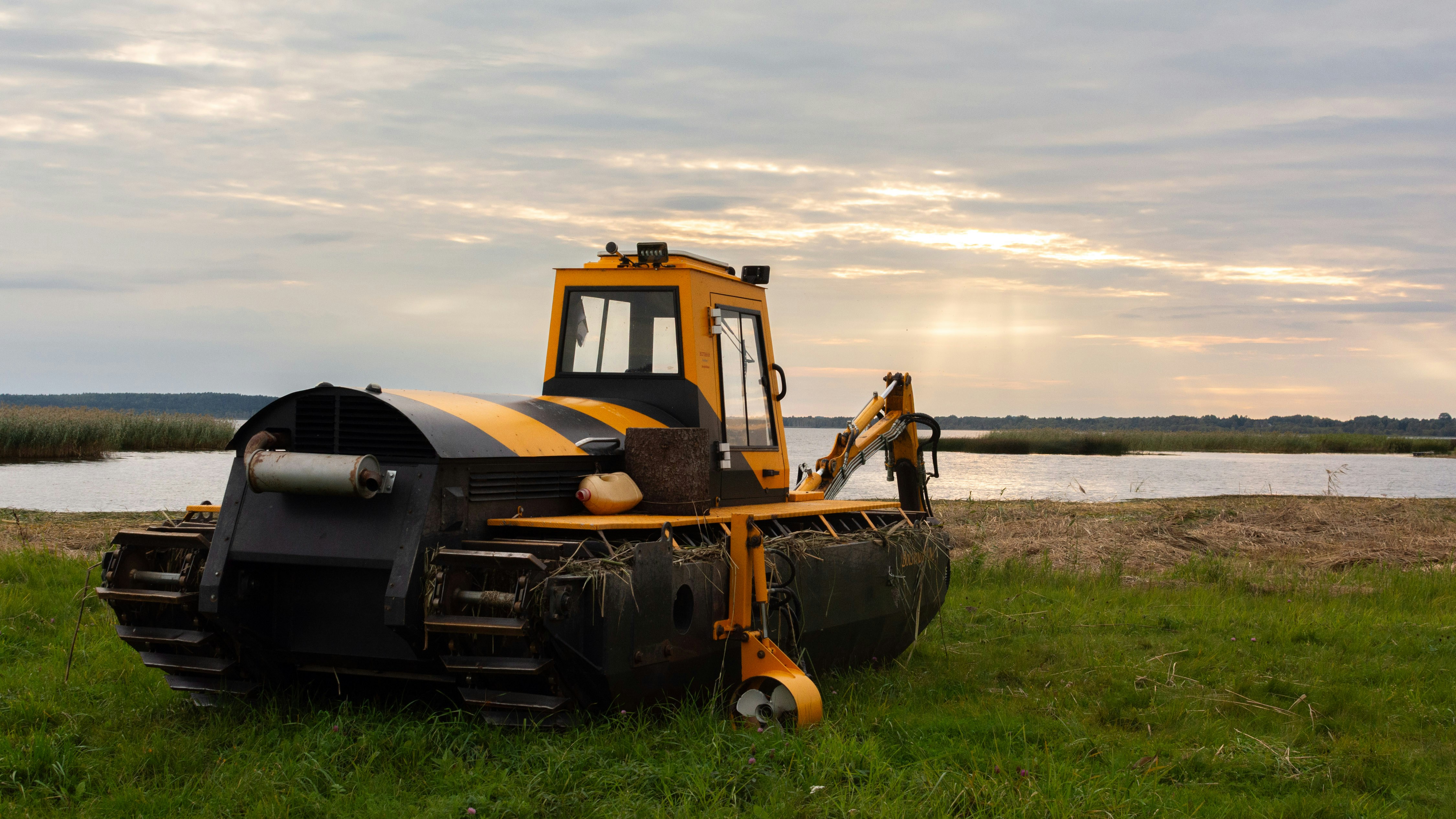 Yellow tracked vehicle sits on grassy riverbank with a calm waterway in the background; muted sunset light highlights the machine's contours. This is a documentary-style landscape shot of a heavy-duty dredger-like unit.