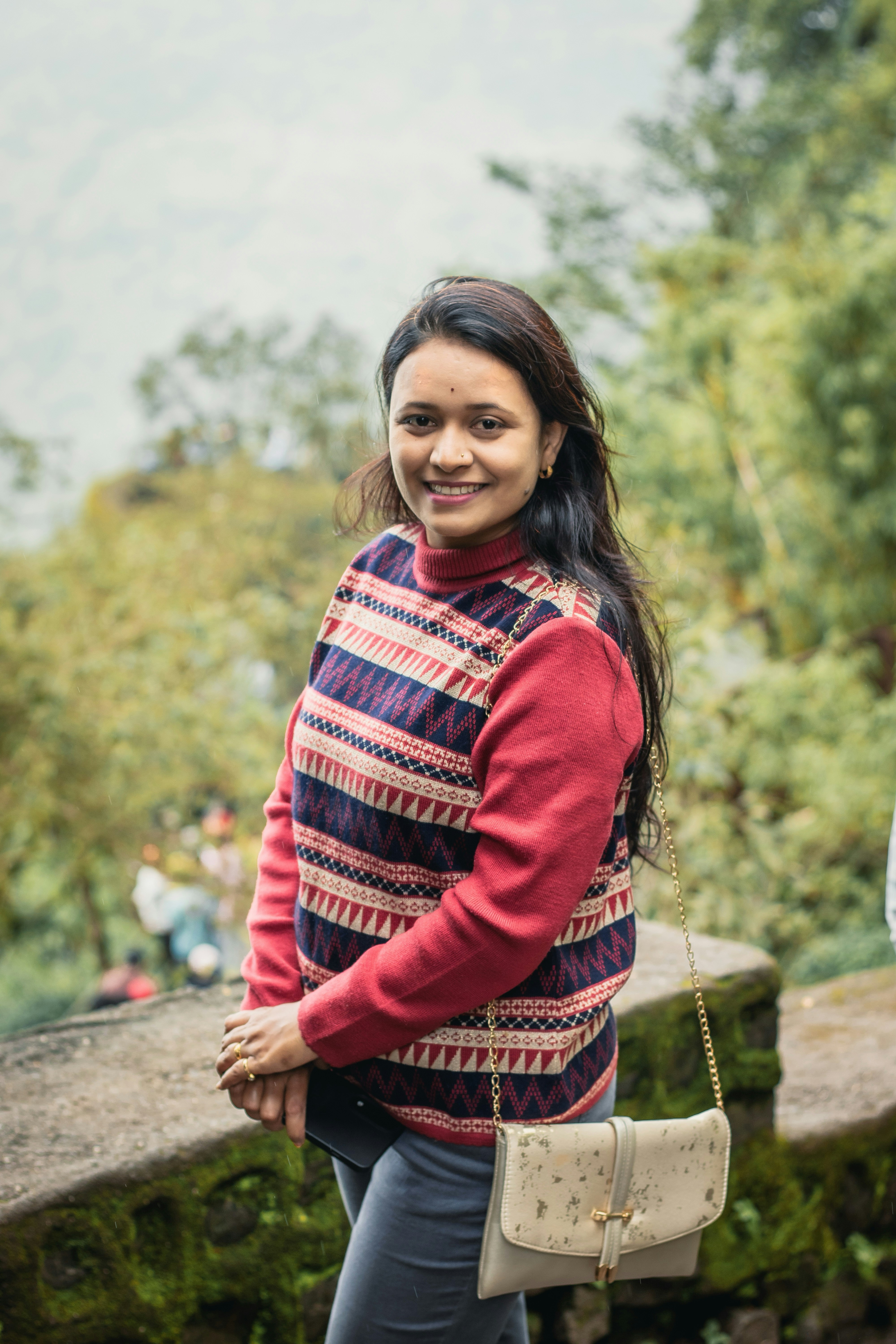 Portrait of a smiling woman in a patterned red sweater standing outdoors with a soft, green background; a beige handbag rests at her side, creating a casual travel vibe.
