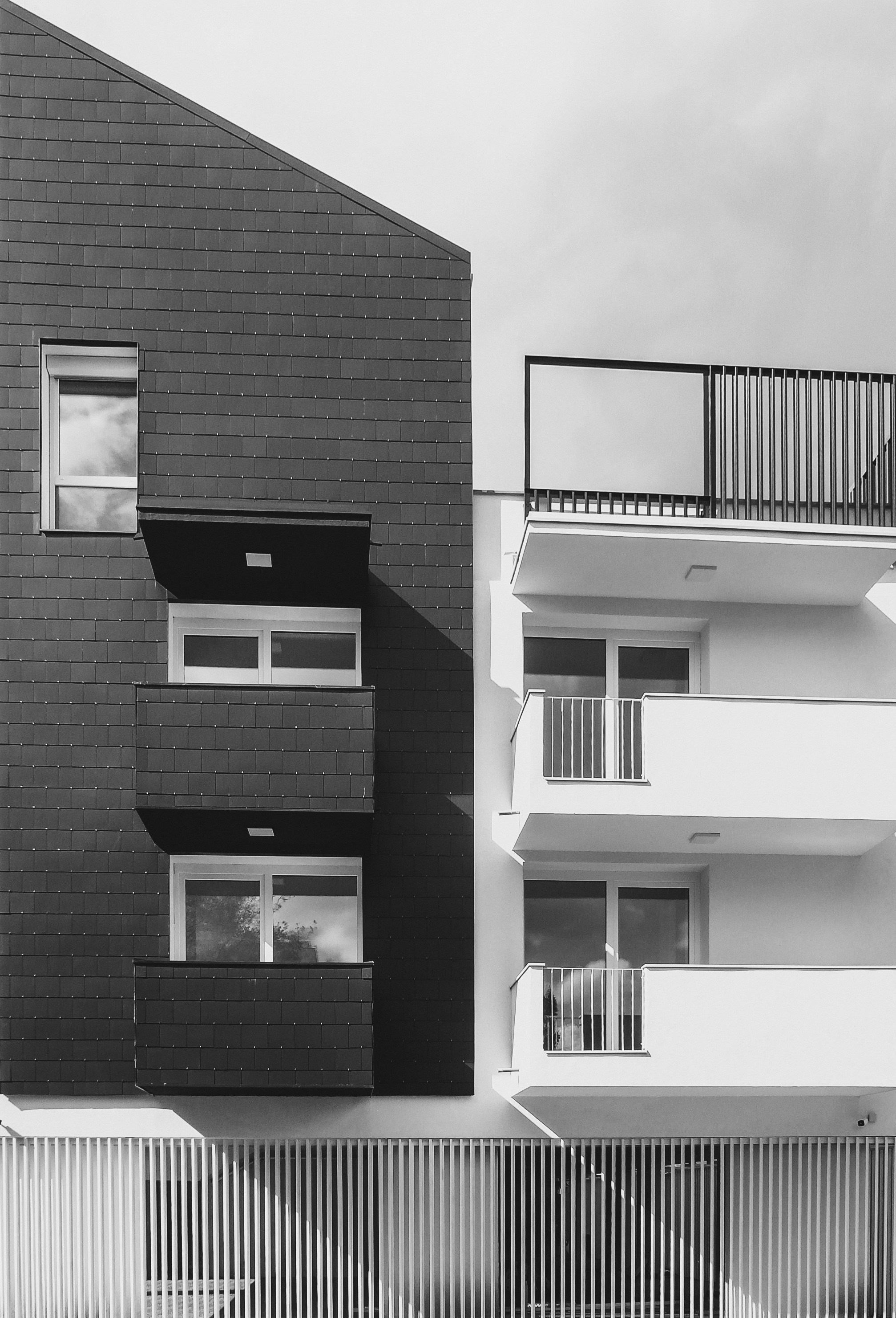 Monochrome shot of a modern building facade with a dark, textured left wing and a white, smooth right wing, featuring balconies and vertical railing elements.