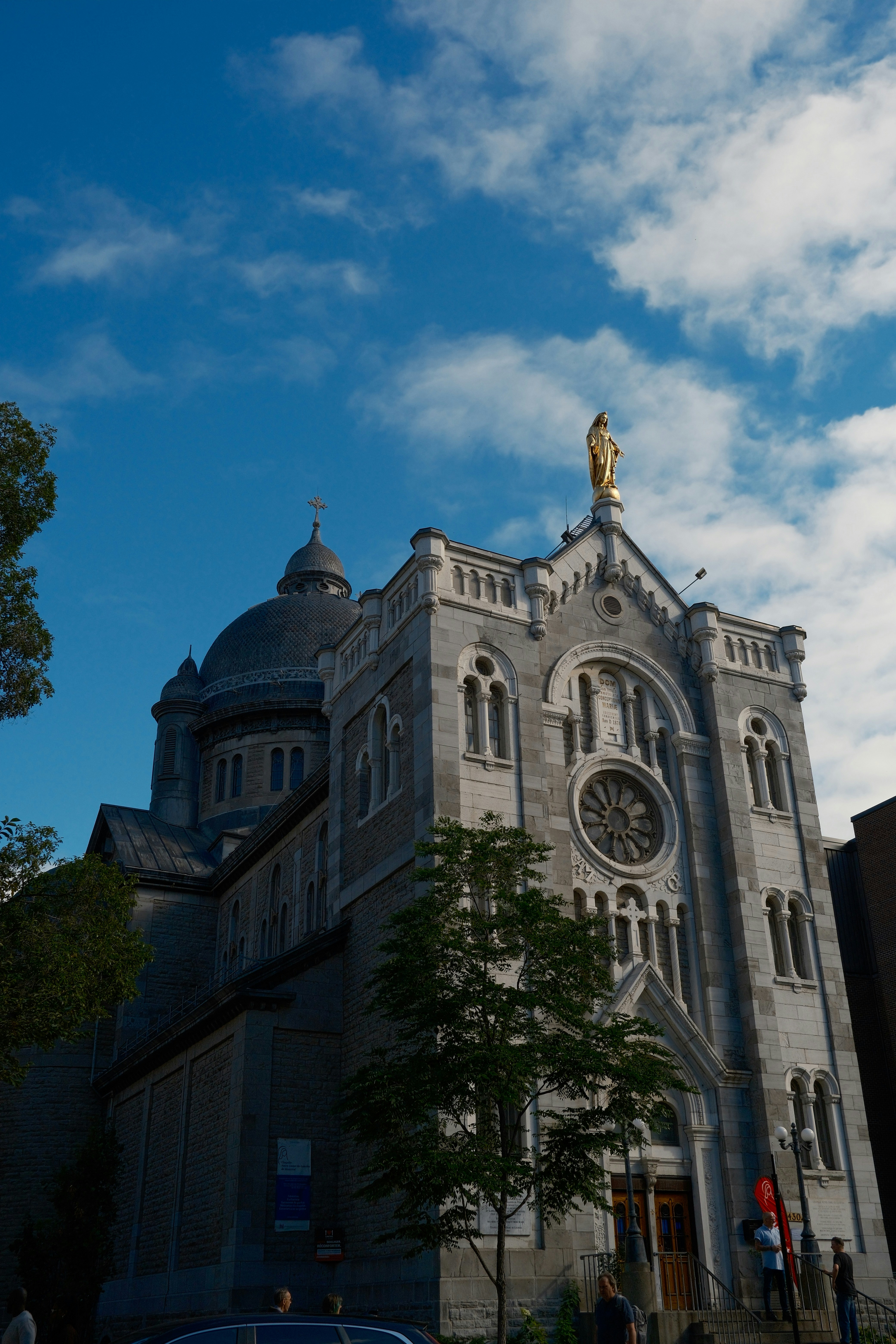 Grey stone cathedral façade with a circular rose window and a golden statue atop, framed by a canopy of trees against a bright blue sky.