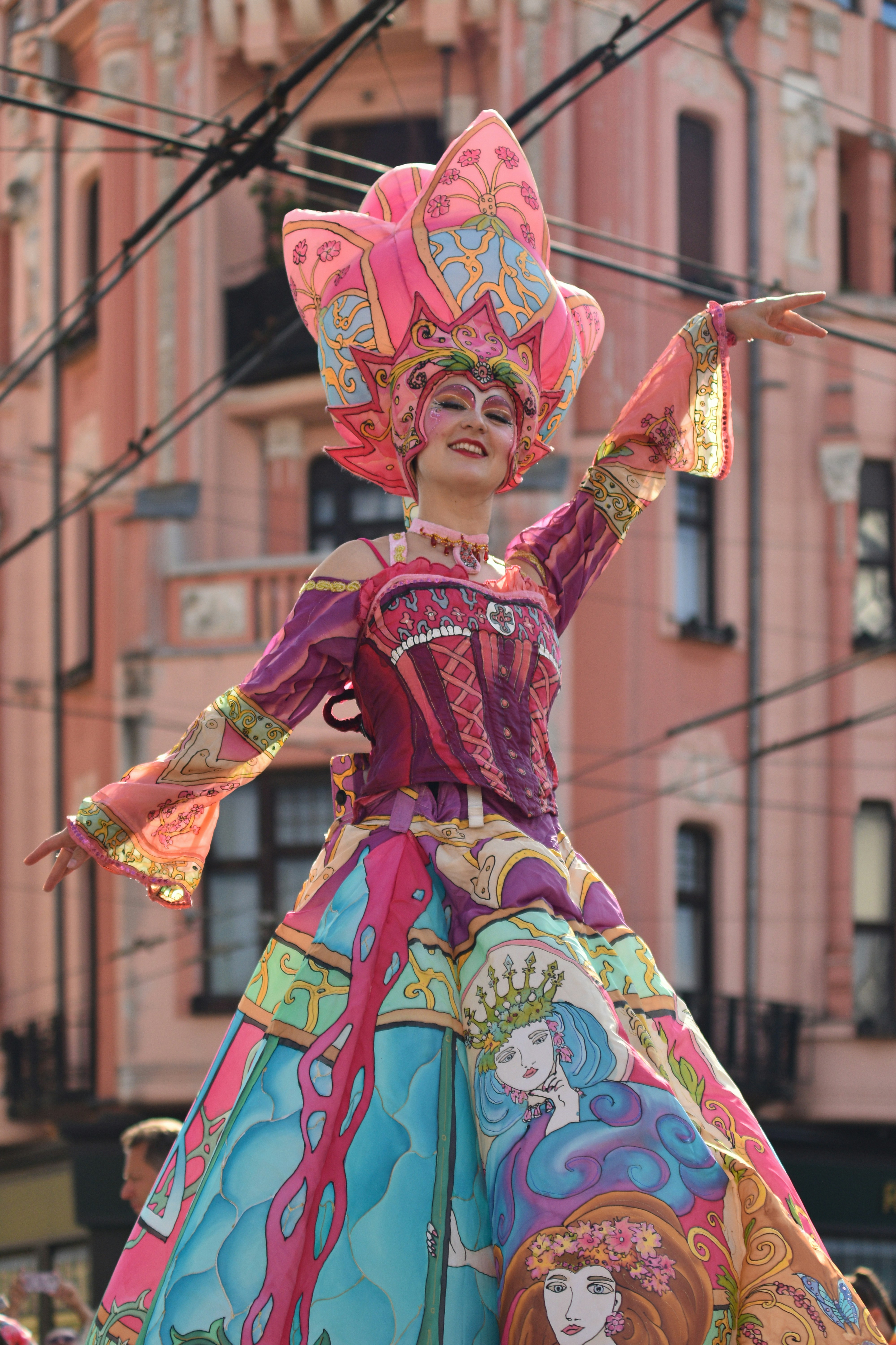 Colorful carnival performer in a decorated pink and turquoise costume with an oversized headpiece, dancing in a city street; the scene includes pink facades and overhead tram cables.