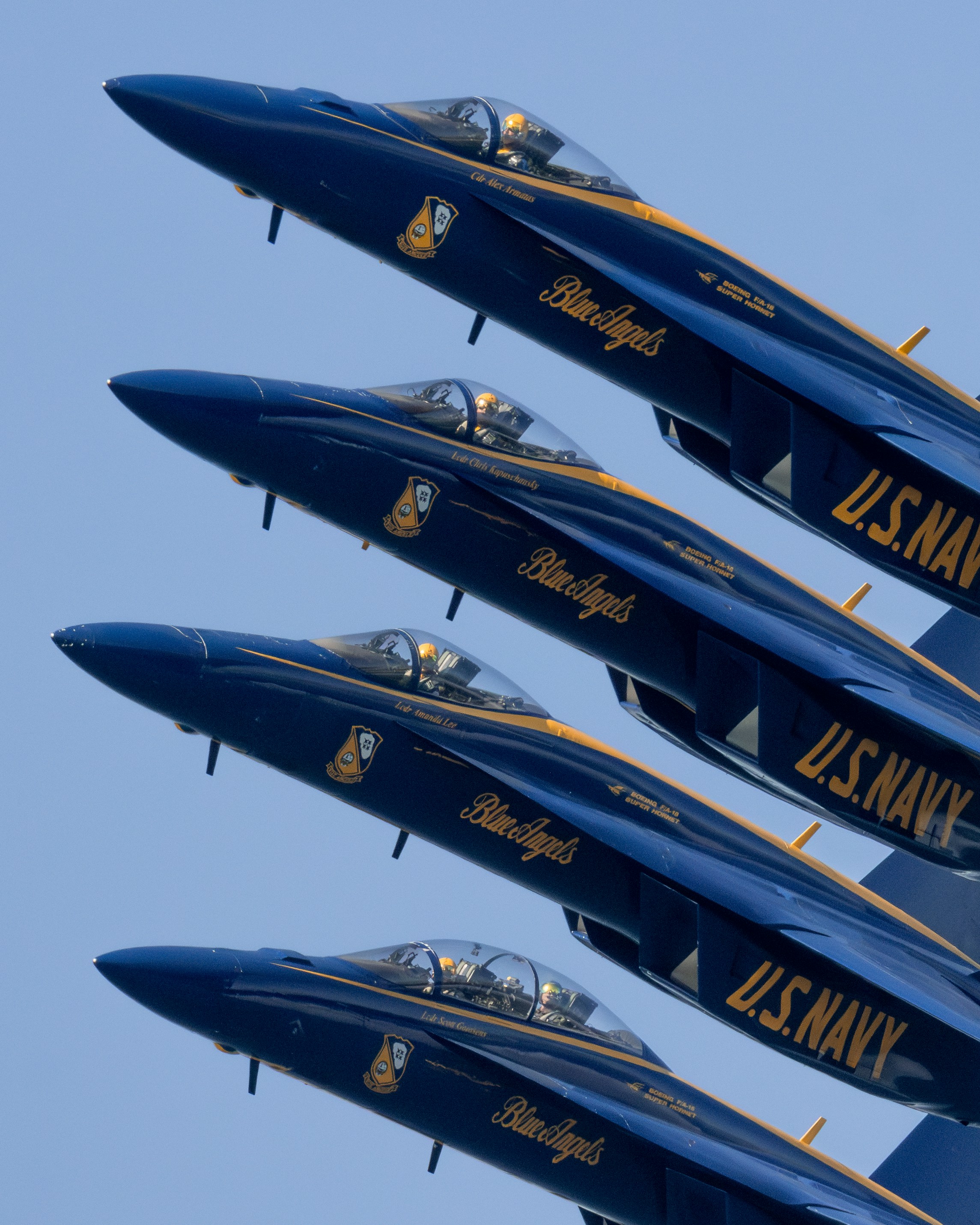 Four blue U.S. Navy jets in tight, diagonal formation against a clear sky, smoke-free and high-contrast blue fuselages with 'Blue Angels' lettering visible.