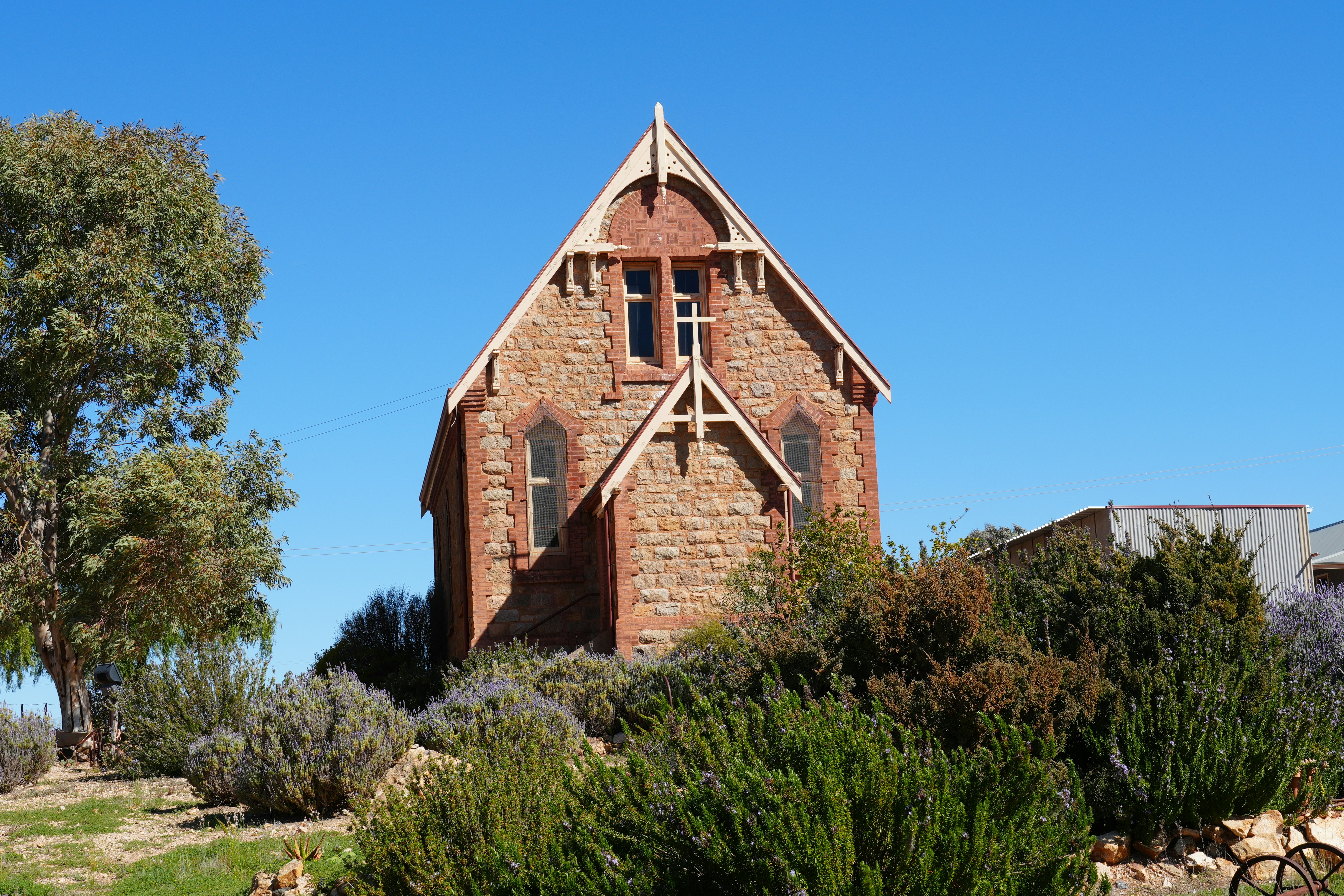A small stone chapel set among greenery and shrubs under a clear blue sky.