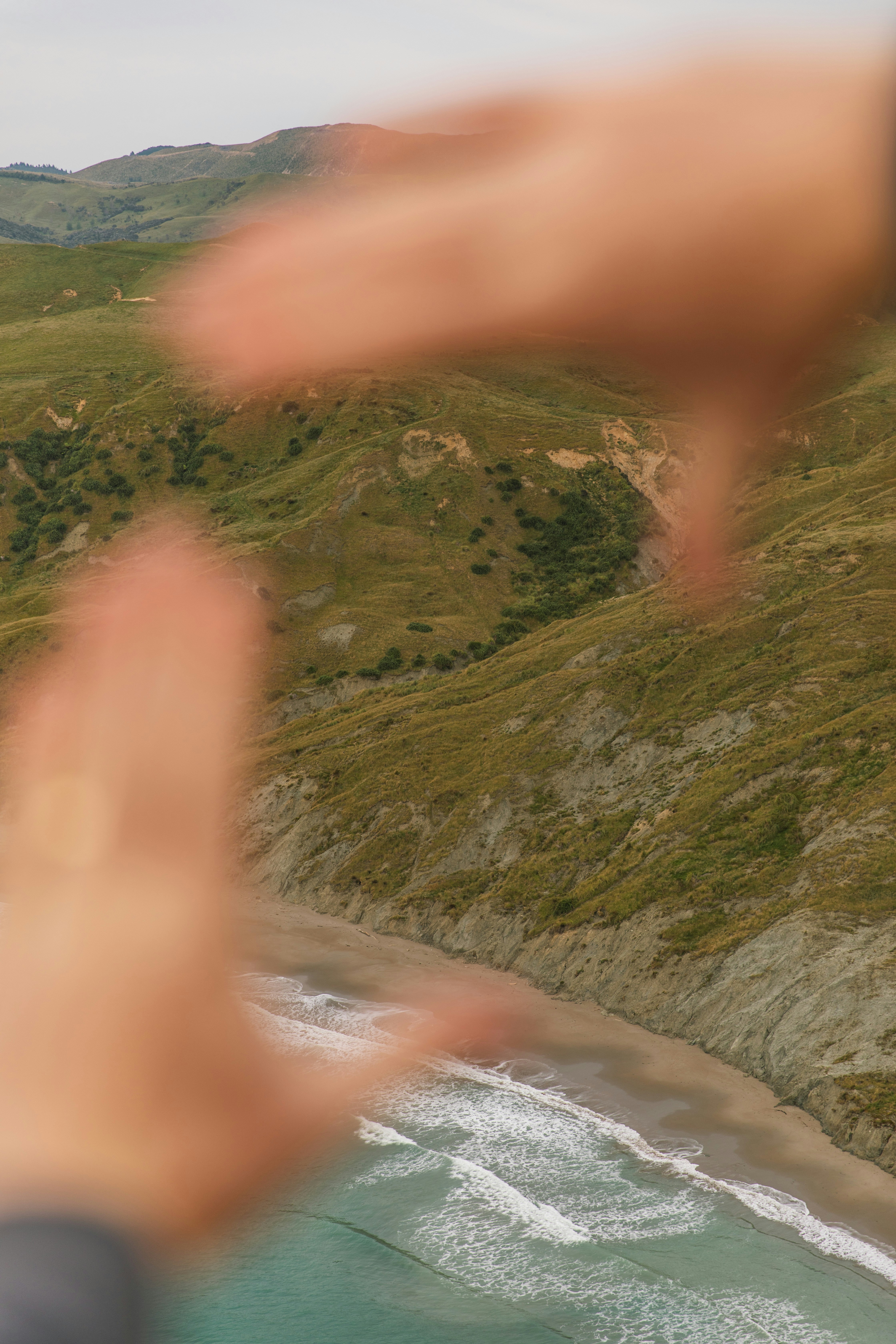 Blurry peach-hand silhouettes frame a sunlit coastal hillside and turquoise shoreline, with the landscape visible through the gaps. The image appears as a conceptual, painterly capture rather than a direct portrait.