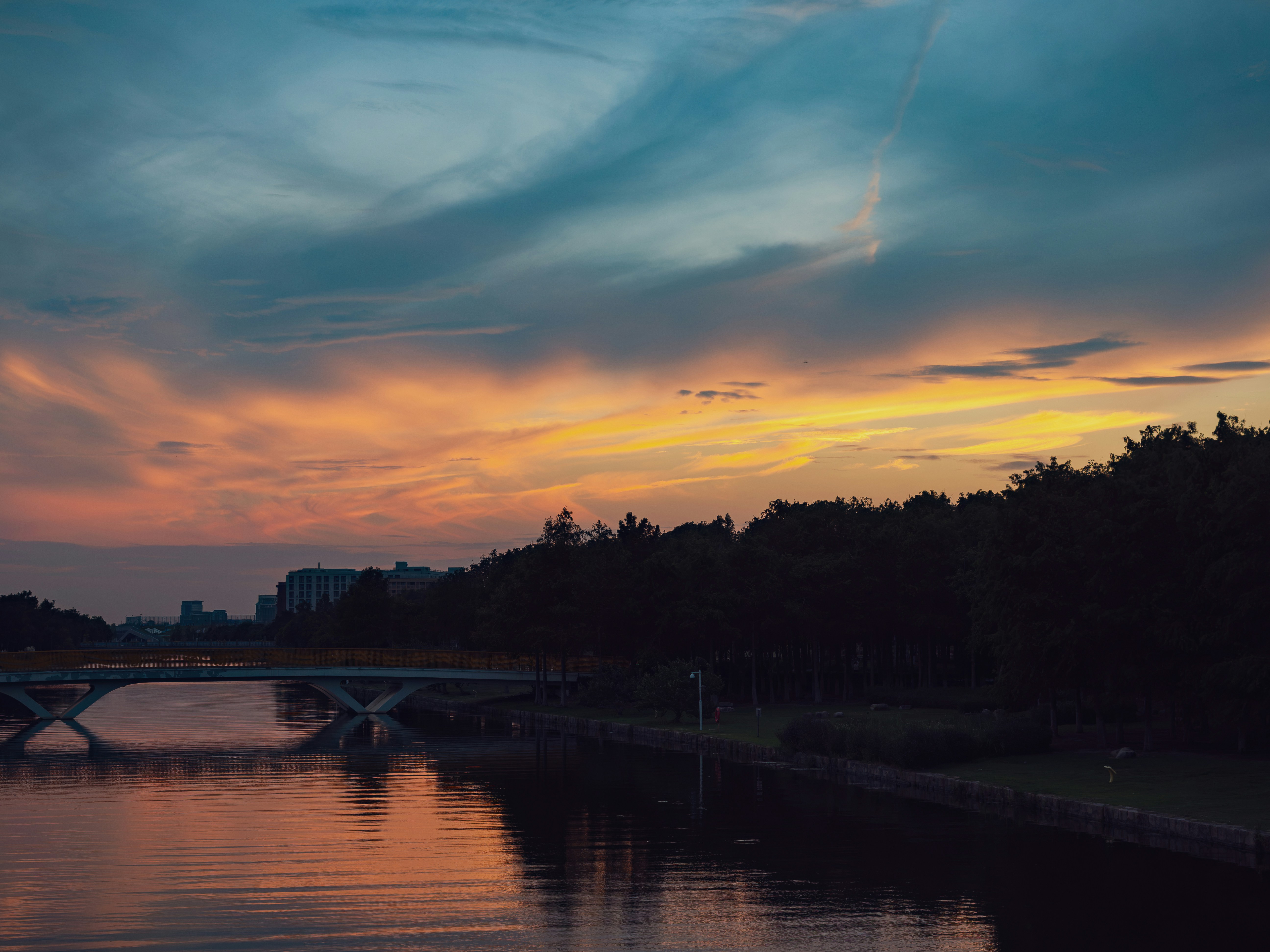 A tranquil riverside scene at sunset with a calm water surface, a distant bridge, and a dark tree line framing the glowing sky. A logo or structure is subtly visible on the left bank.