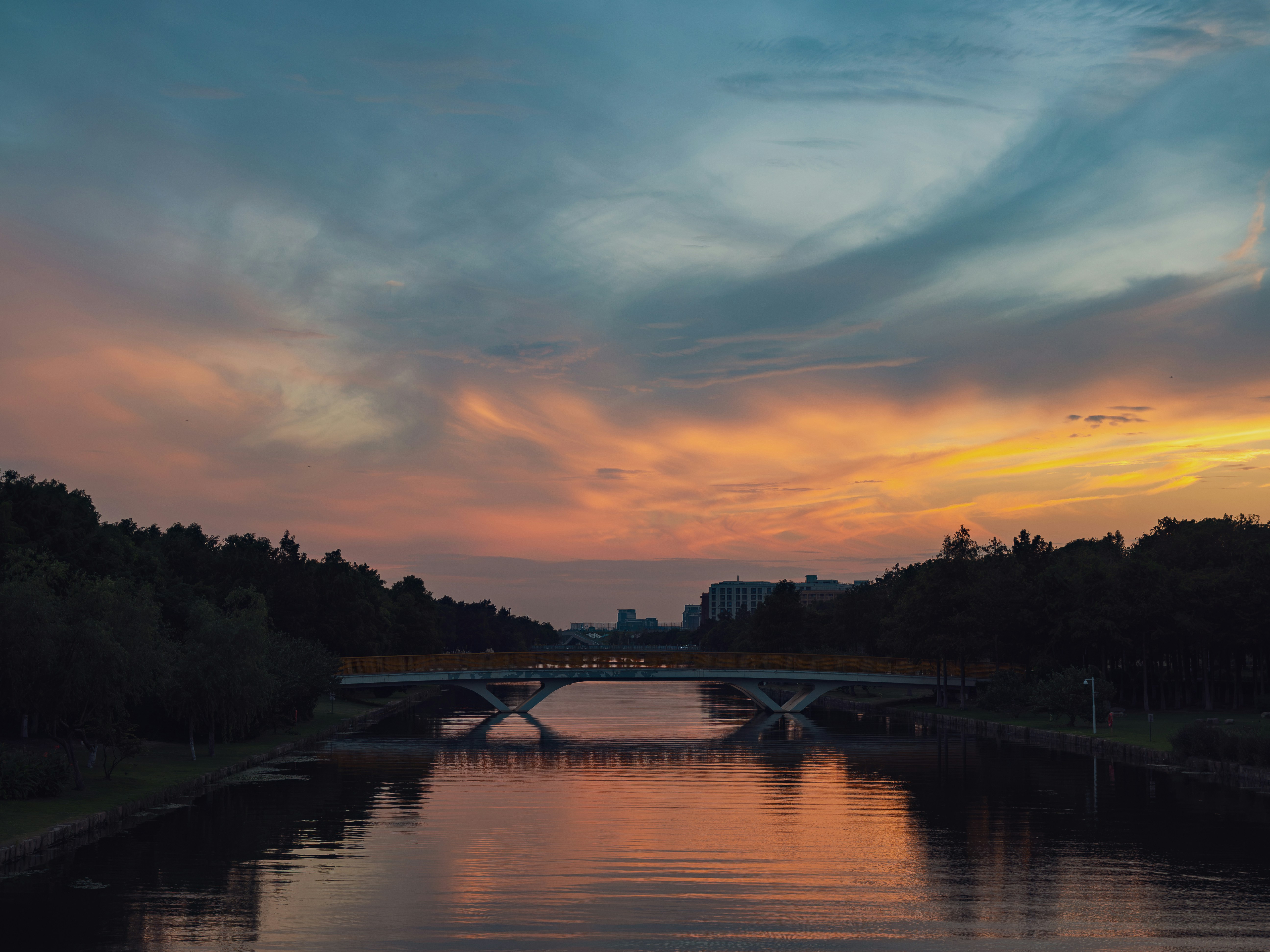 Amber and pink clouds reflect on a calm river flanked by trees, with a pedestrian bridge stretching across the water at dusk.