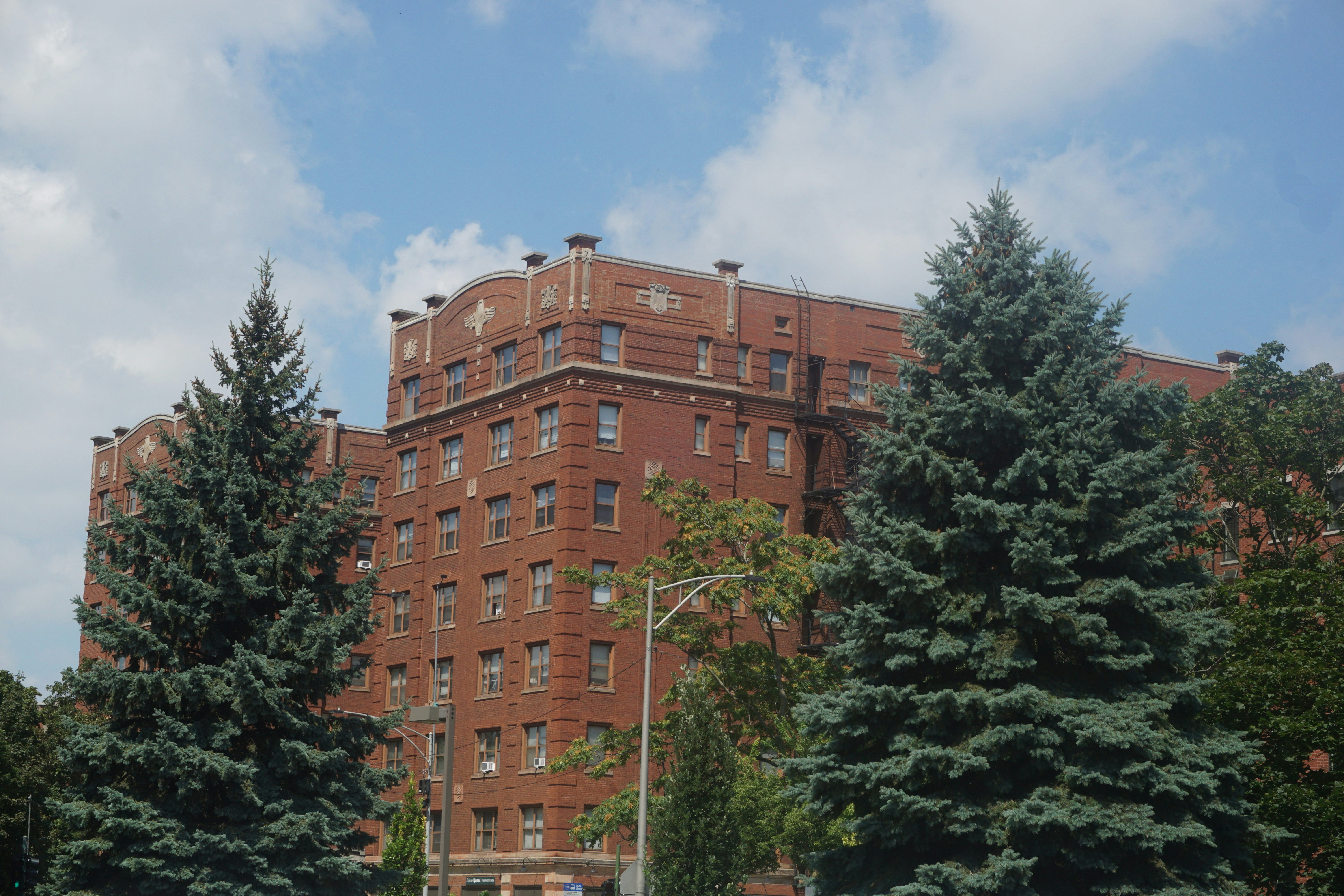 Brick apartment building framed by evergreen trees under a bright, partly cloudy sky. A streetlamp rises in front, hinting at urban life around the historic facade.