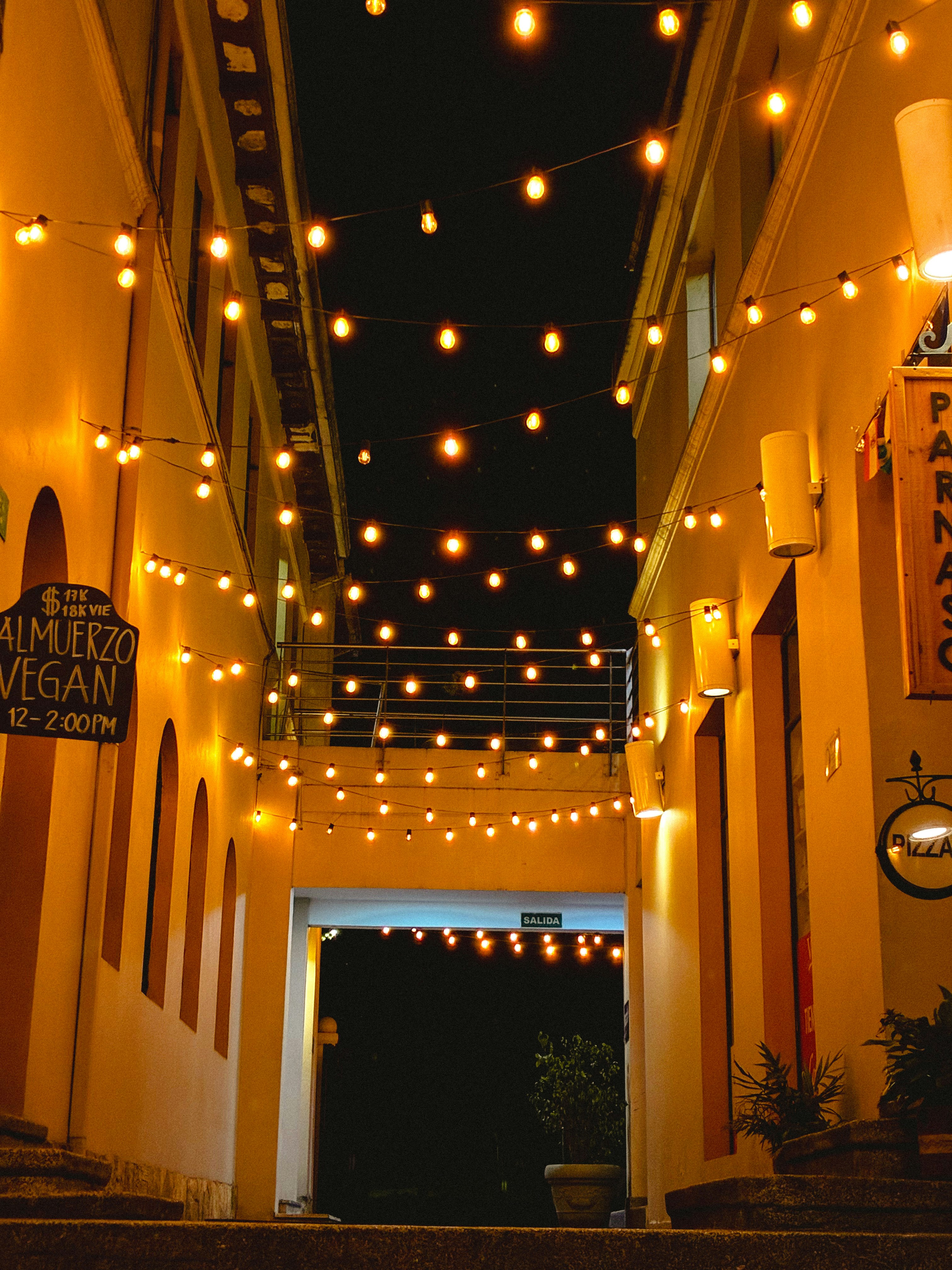 Warm string lights crisscross a narrow alley framed by yellow walls, guiding the viewer toward an arched exit at dusk and a dark door beyond. A subtle vegan sign and potted plants hint at a cozy, urban dining alley.