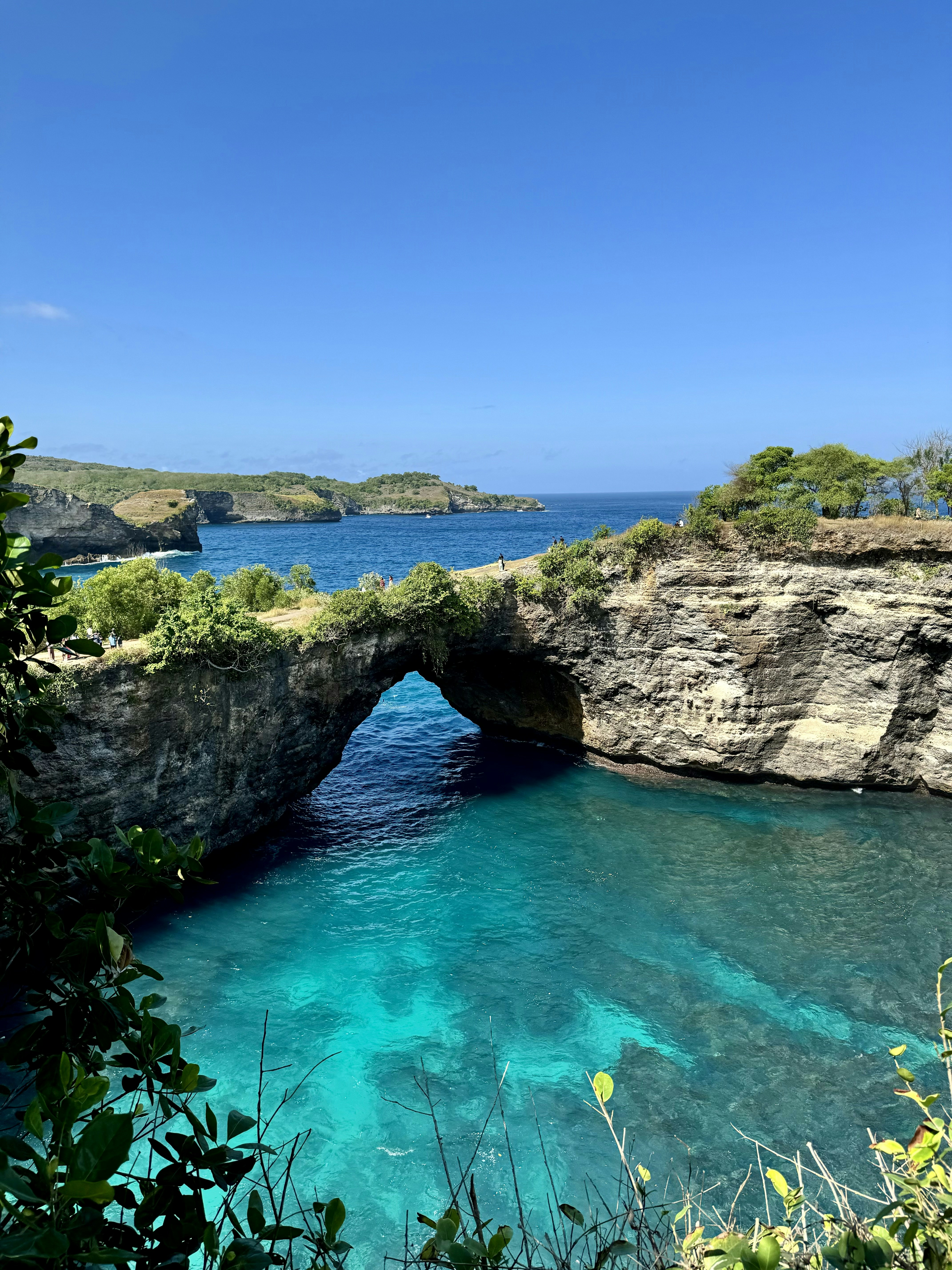 Vivid turquoise water flows beneath a natural rock arch along a rugged coastline, with blue sea and sky in the background. A natural coastal landscape showcasing clear water and sunlight.